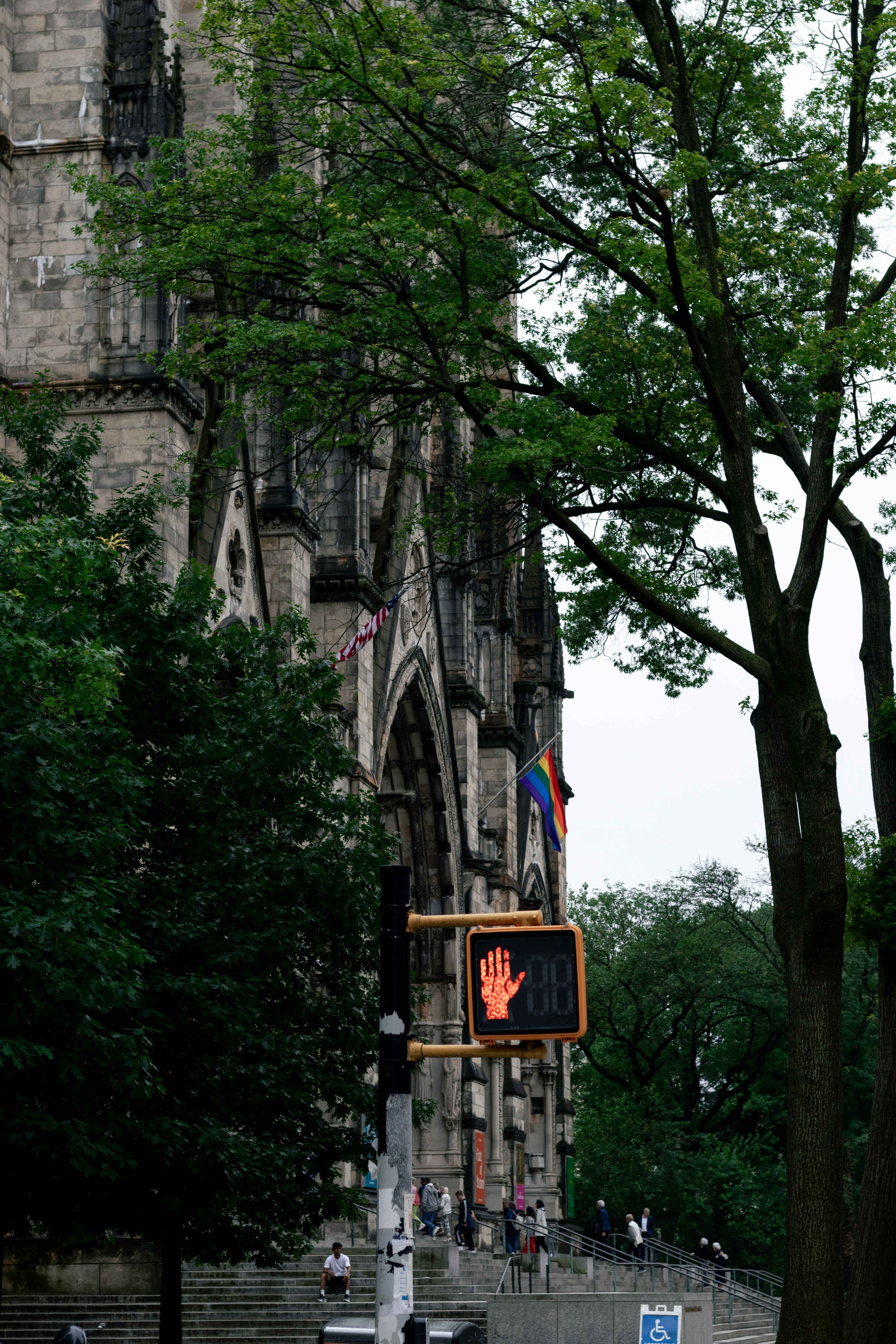 Traffic signal displaying a hand symbol against a backdrop of an architectural structure and lush greenery.