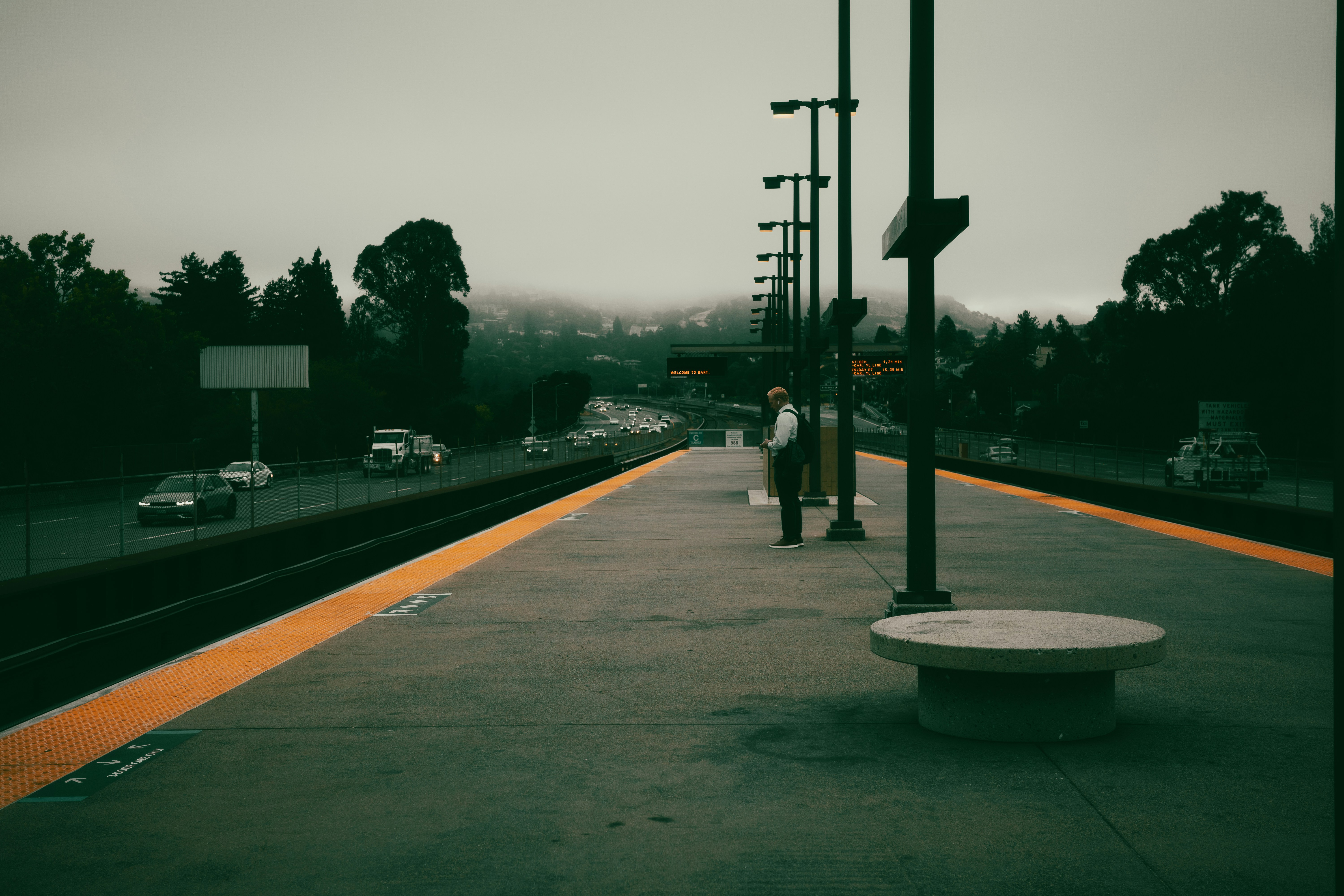 A lone figure stands on a train platform, surrounded by a misty atmosphere and distant traffic. The scene captures the anticipation of travel amid a muted landscape.