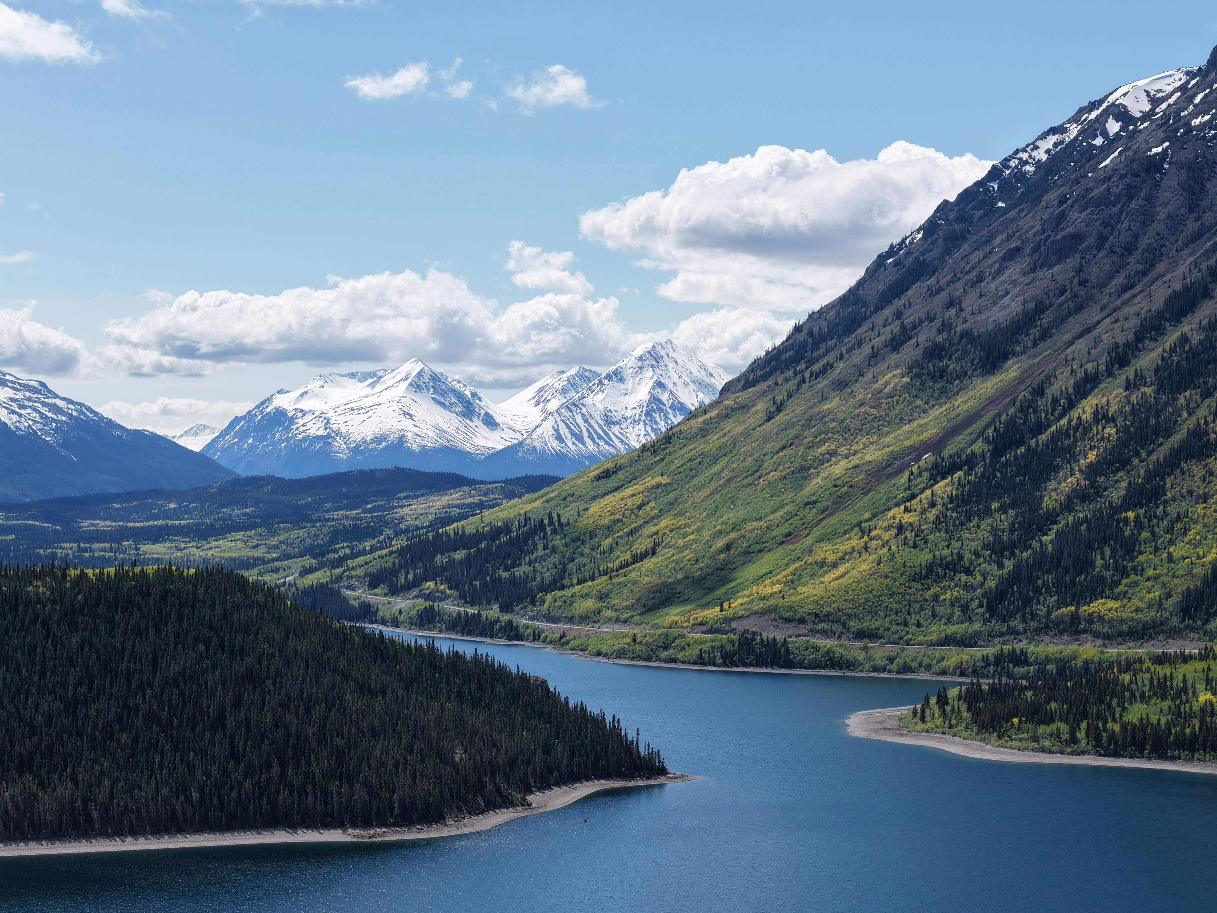 Lush green hills gently slope down to a winding river, with snow-capped mountains rising majestically in the background under a partly cloudy sky.