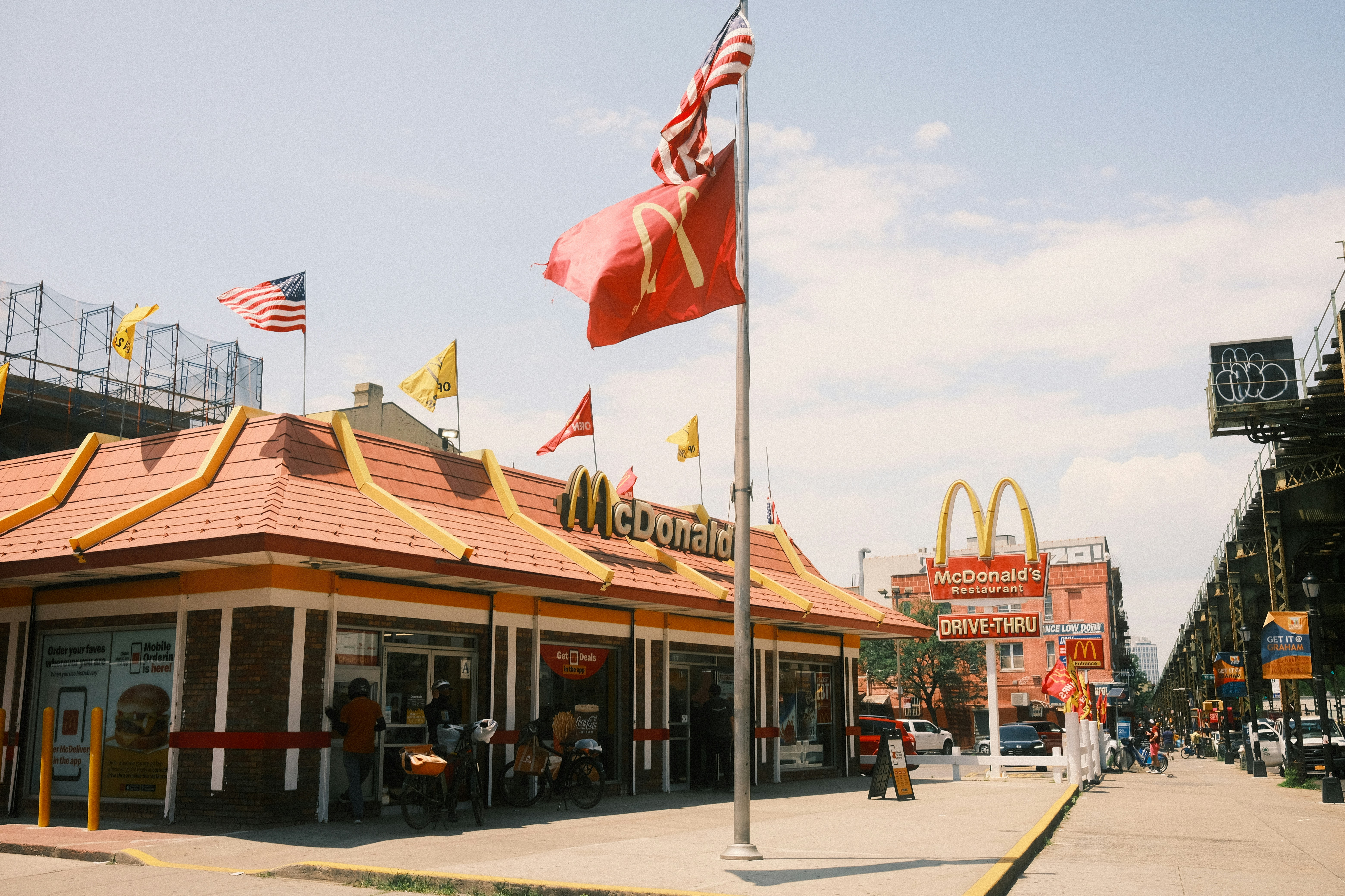 McDonald's restaurant with vibrant flags and signage, set against a bustling city backdrop. Drive-thru lane visible on the right.