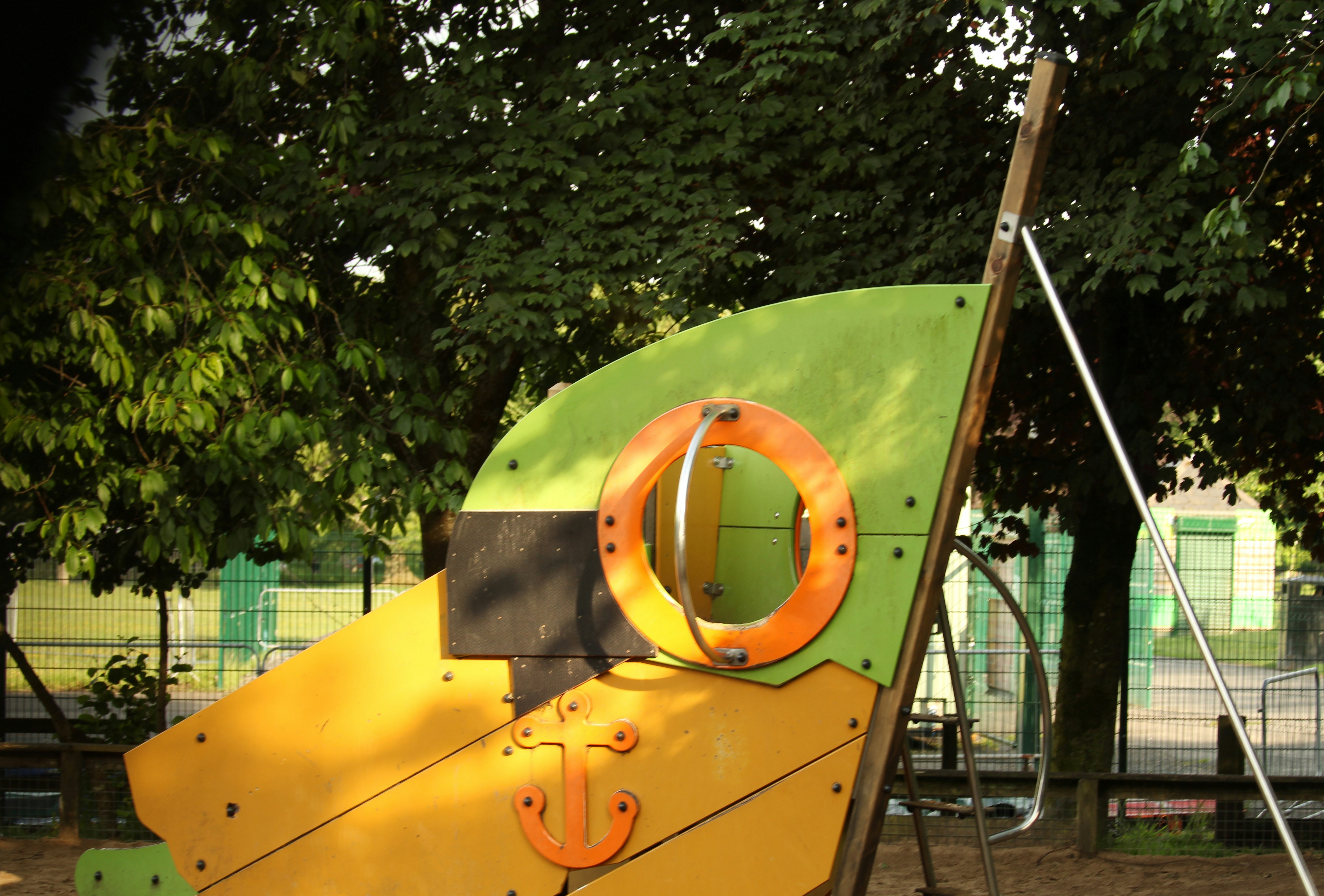 Colorful playground structure resembling a ship's bow with a porthole and anchor detail, surrounded by greenery.