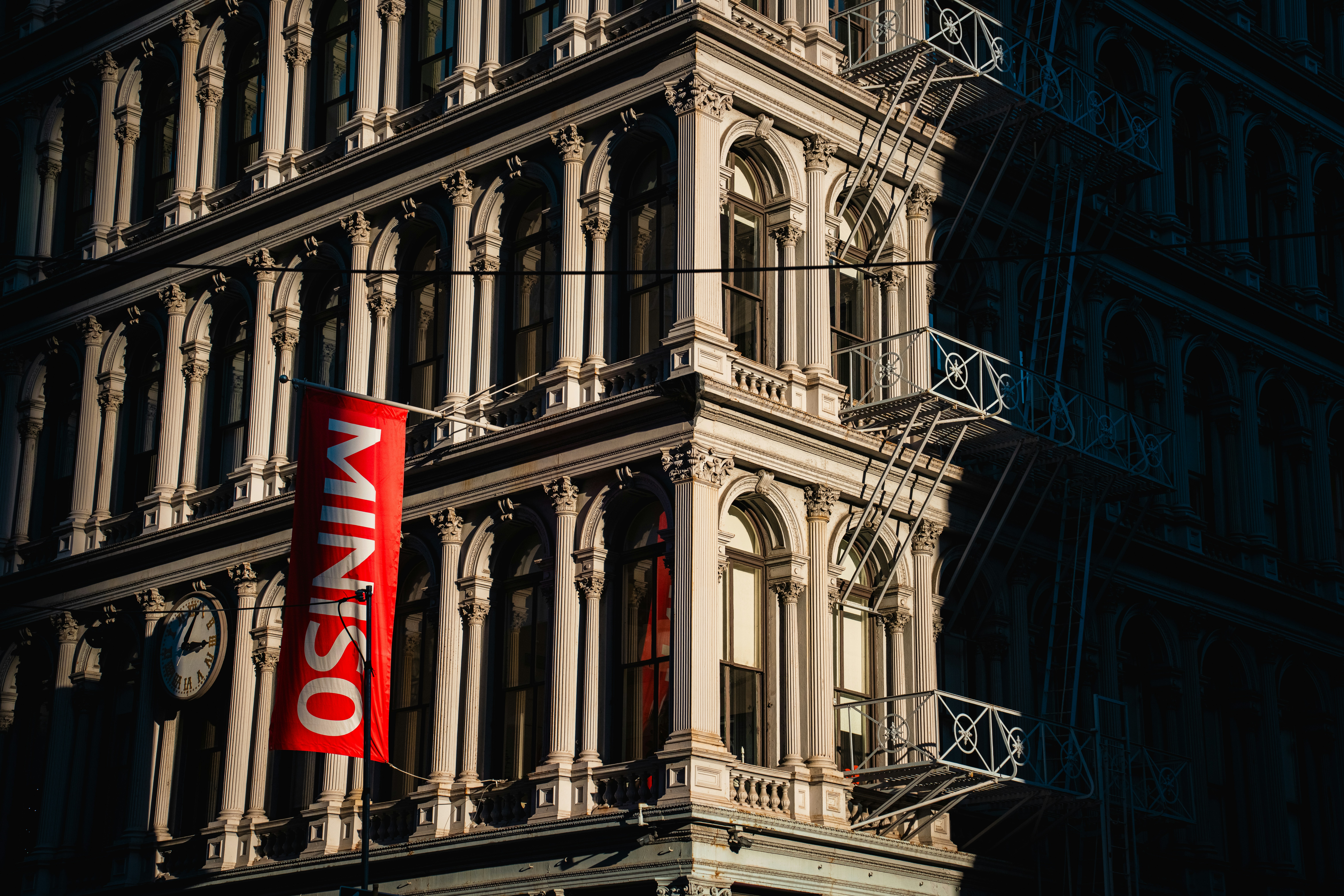 A detailed view of a historic cast-iron building in Soho, New York City, featuring elegant columns, arched windows, and a classic fire escape. Sunlight highlights the architectural details, while a bold red Miniso banner and a vintage clock add pops of color and context to this iconic city corner, blending history with modern retail.