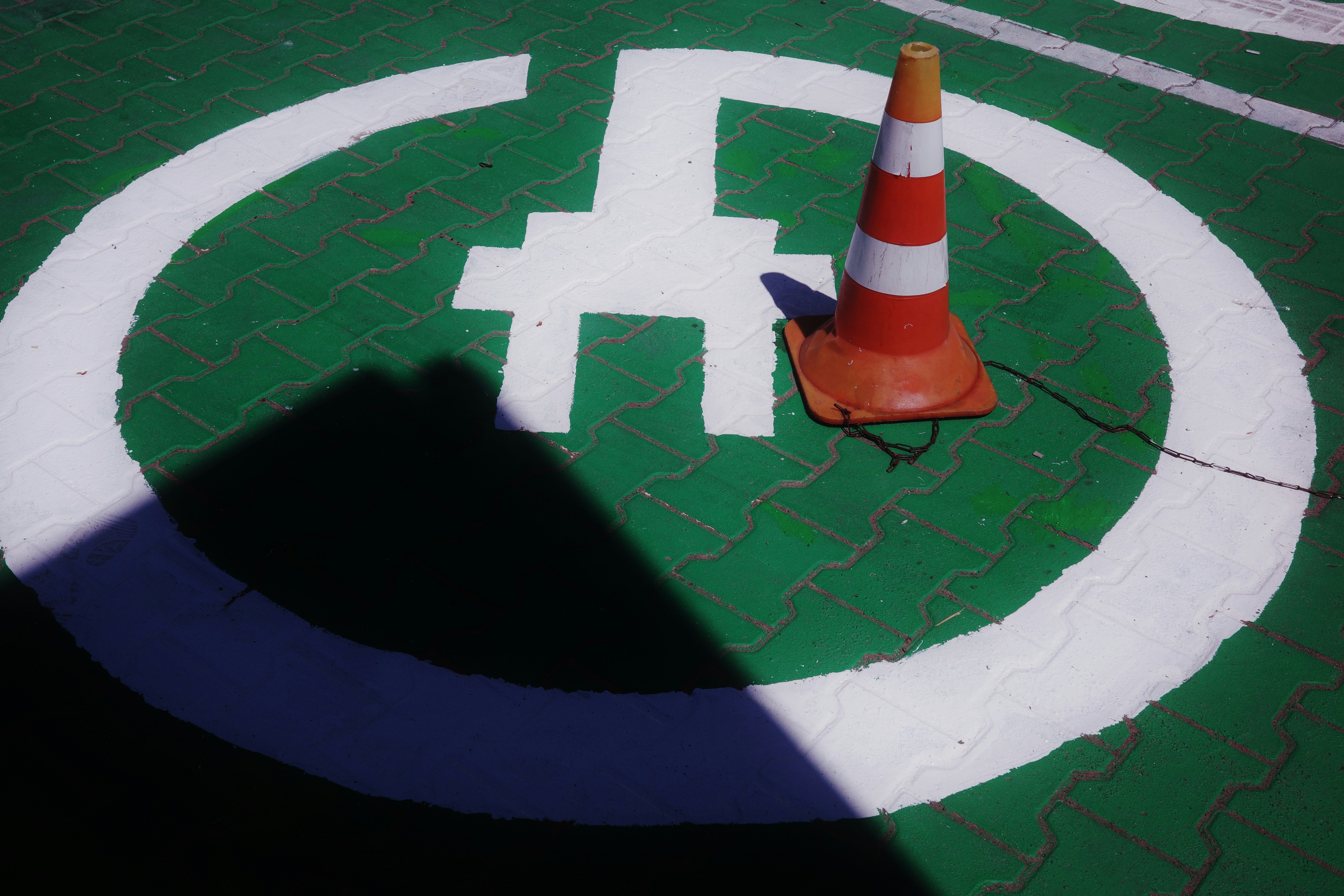 A red and white striped traffic cone sits off-center on a bright green, tiled surface marked with a large white circle and a stylized white 'H' symbol, commonly used for helicopter landing pads. A prominent dark shadow of an unseen object falls across the lower left portion of the marked area, adding depth to the scene. The texture of the interlocking green tiles is clearly visible under the bright light.