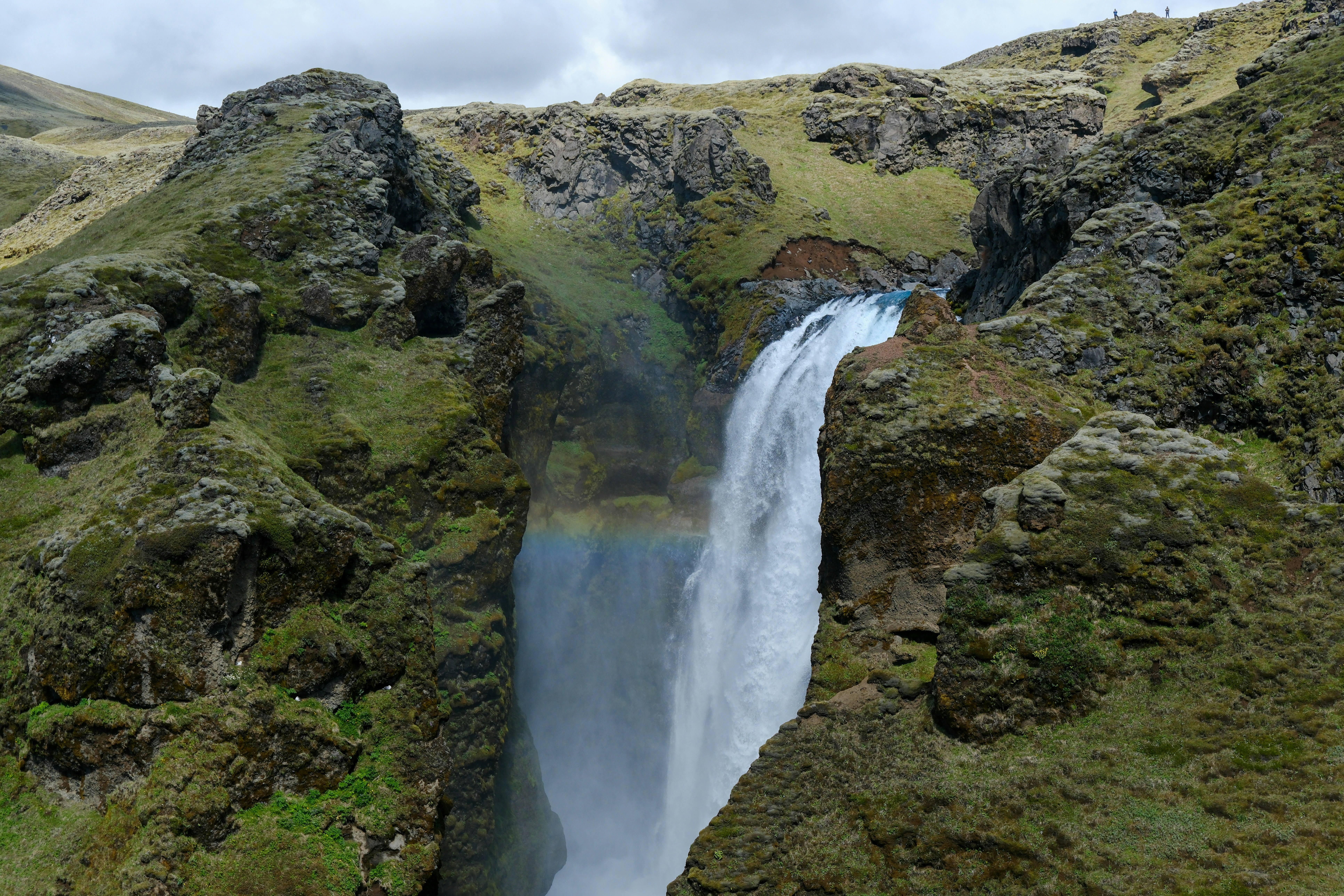 Majestic waterfall cascading down rocky cliffs, surrounded by lush greenery and rugged terrain. A hint of a rainbow emerges from the mist.