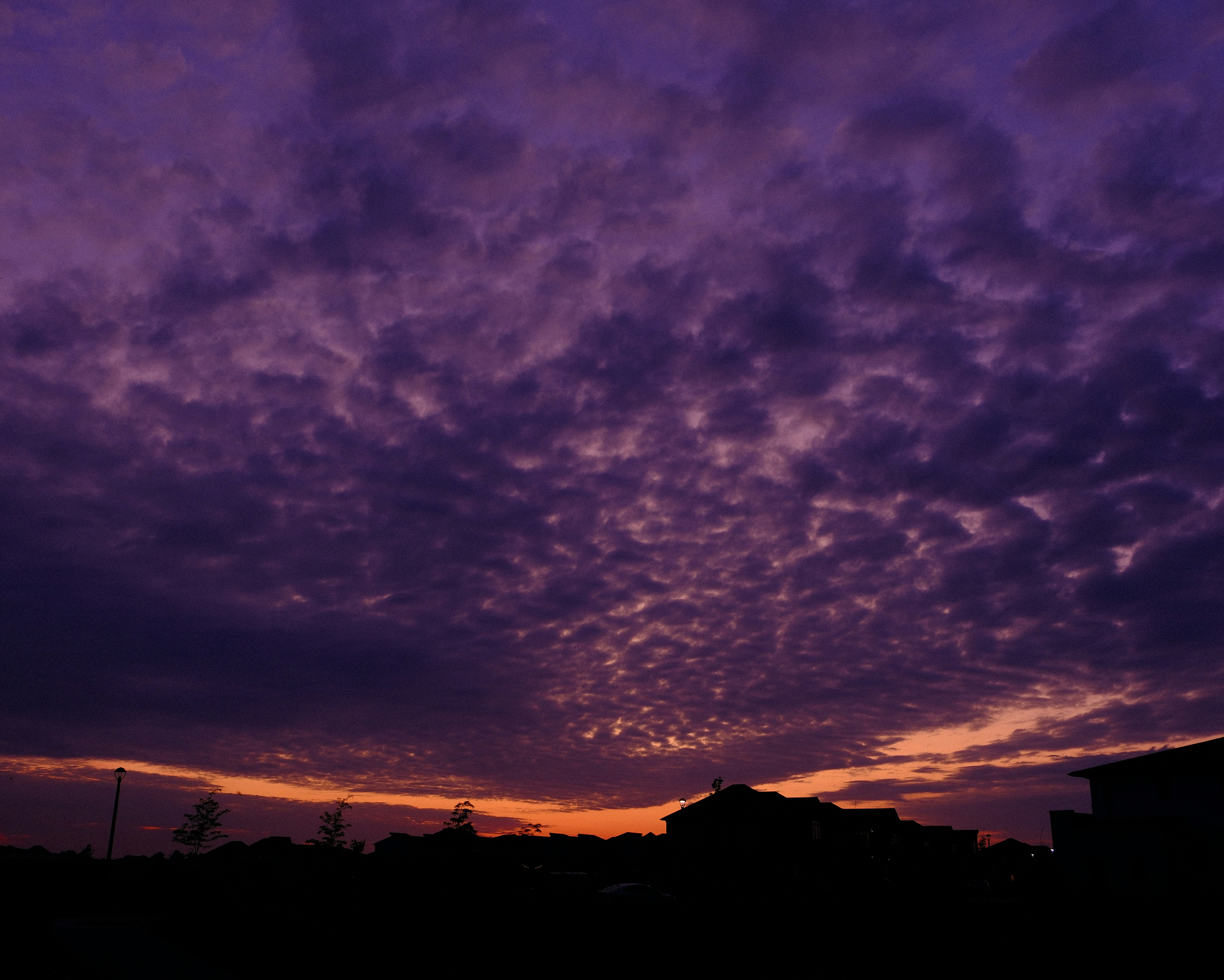 Vibrant twilight sky filled with textured clouds, transitioning from deep purple to warm orange hues, silhouetting distant buildings. 