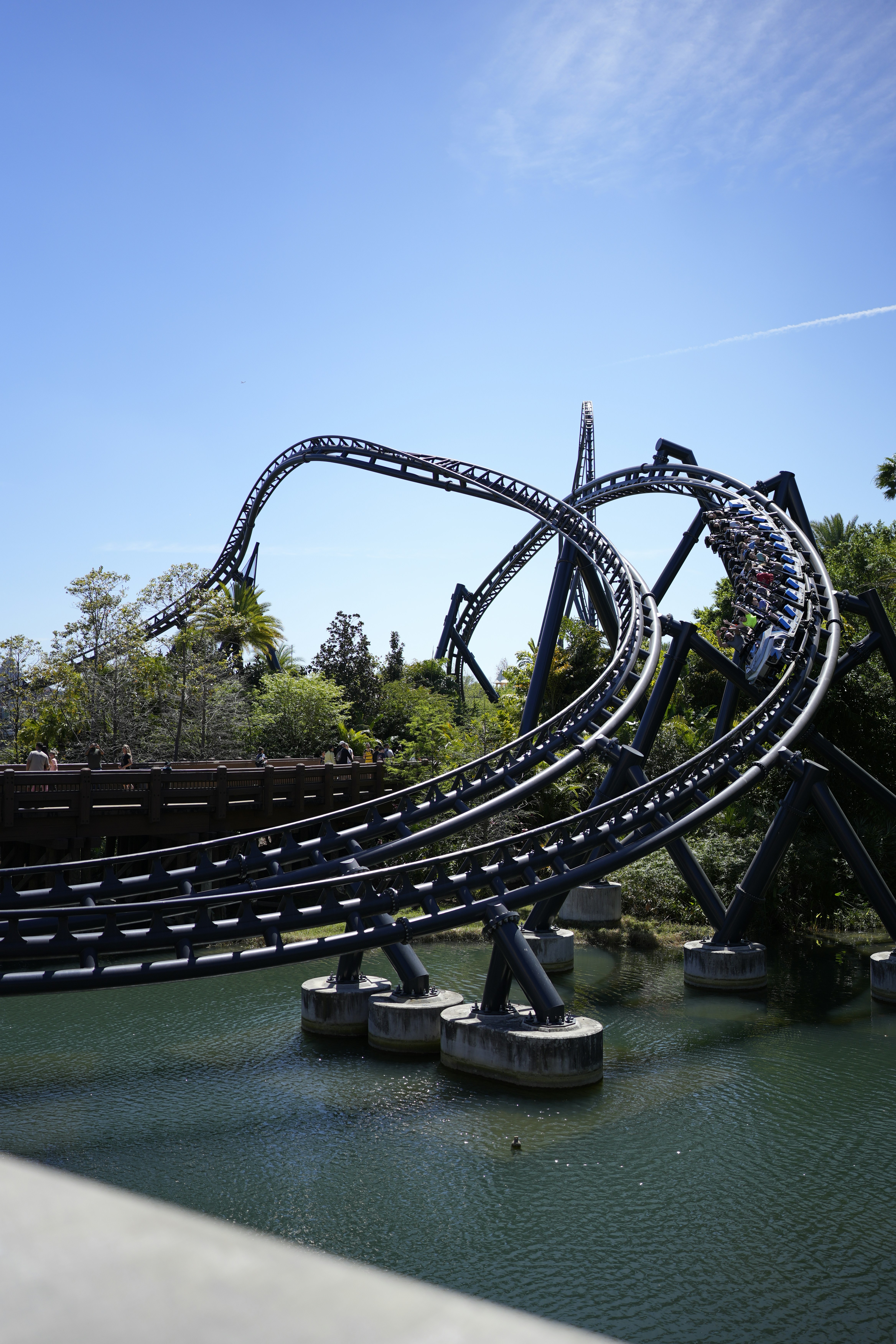 A roller coaster spirals dramatically over a serene body of water, surrounded by lush greenery and clear blue skies.