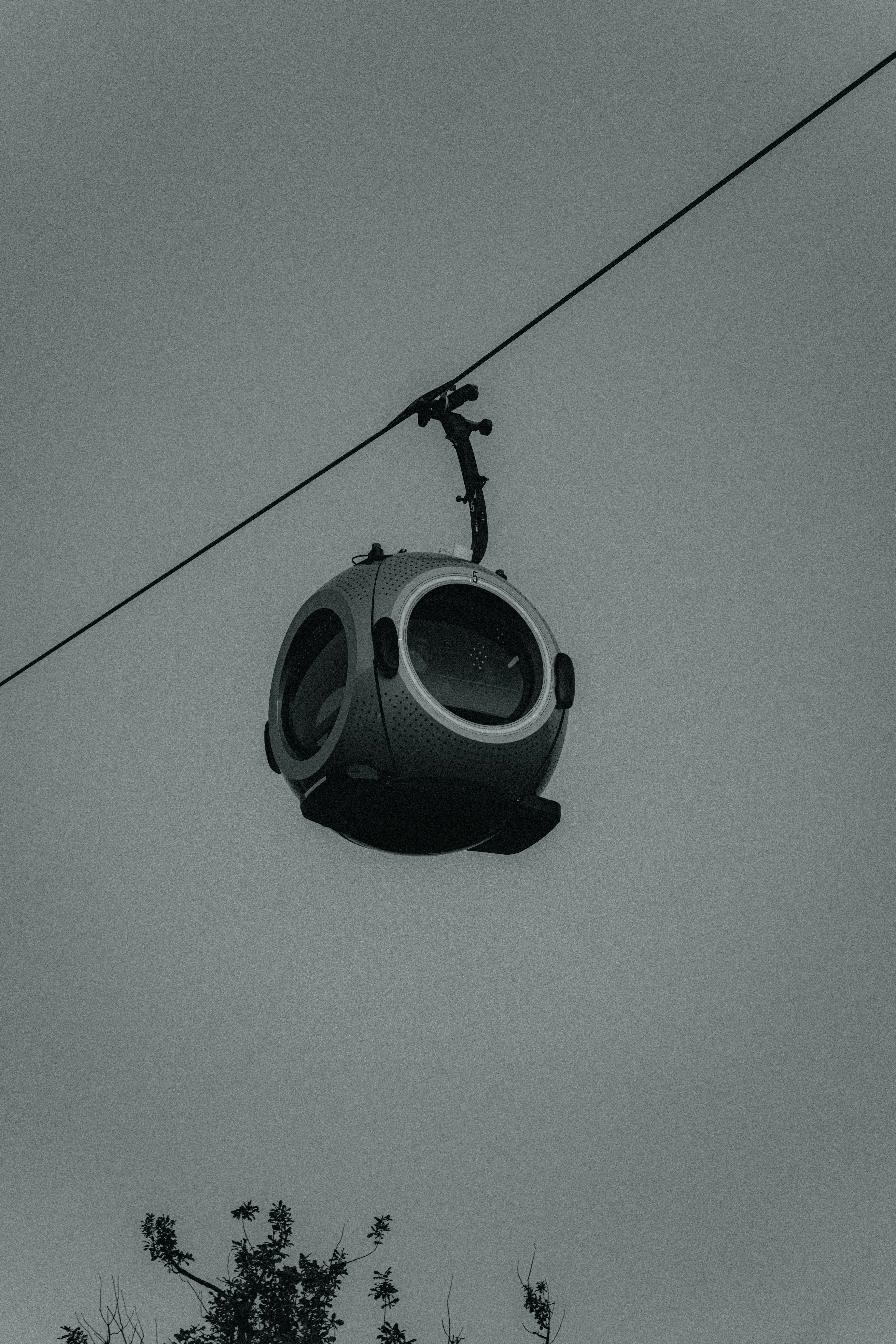A sleek, spherical gondola suspended from a cable against a muted sky, surrounded by silhouetted trees. The design emphasizes modern transportation aesthetics.