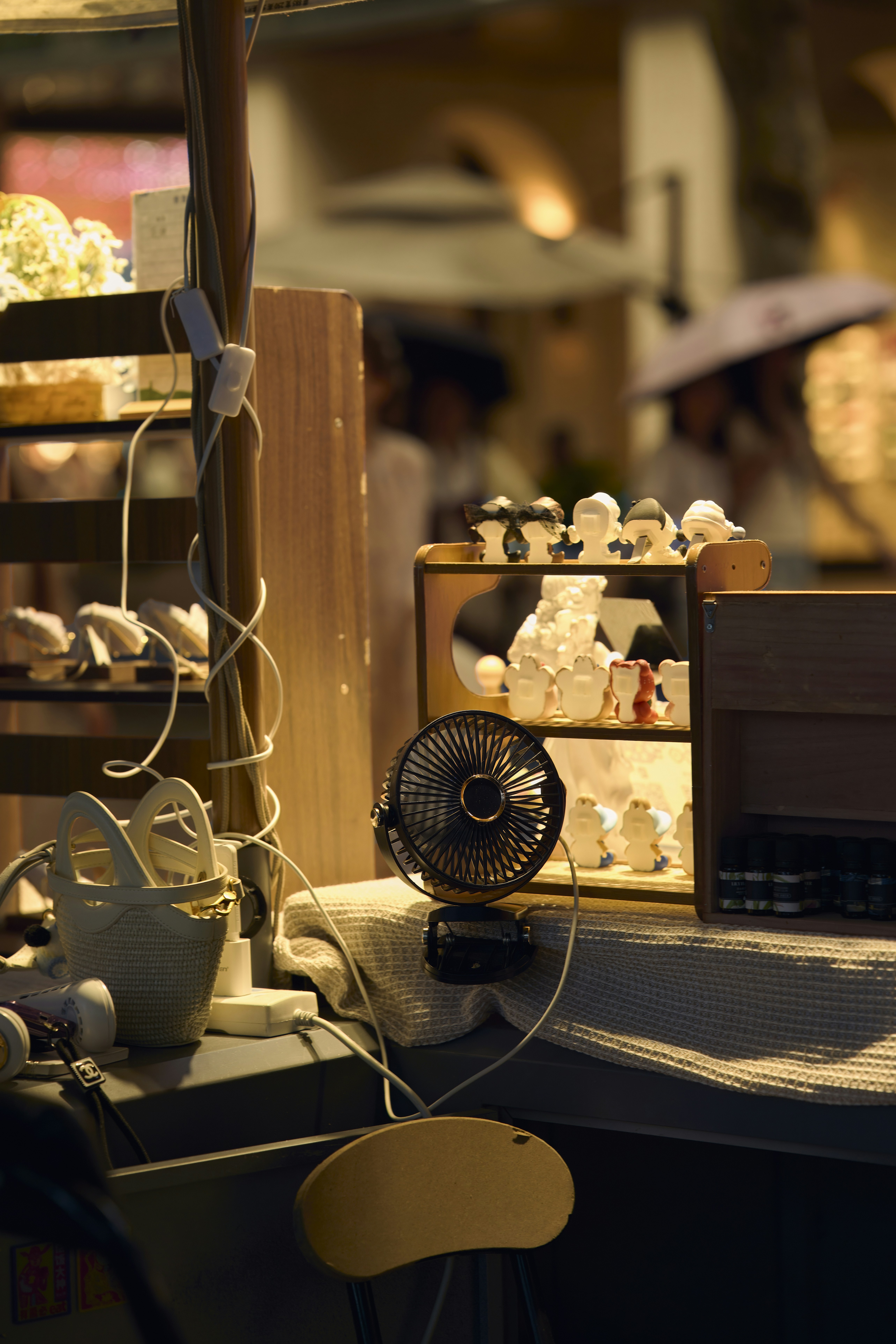 Artisan figurines displayed on a wooden shelf in a bustling market, accompanied by a small fan and a woven bag. Soft lighting enhances the atmosphere.