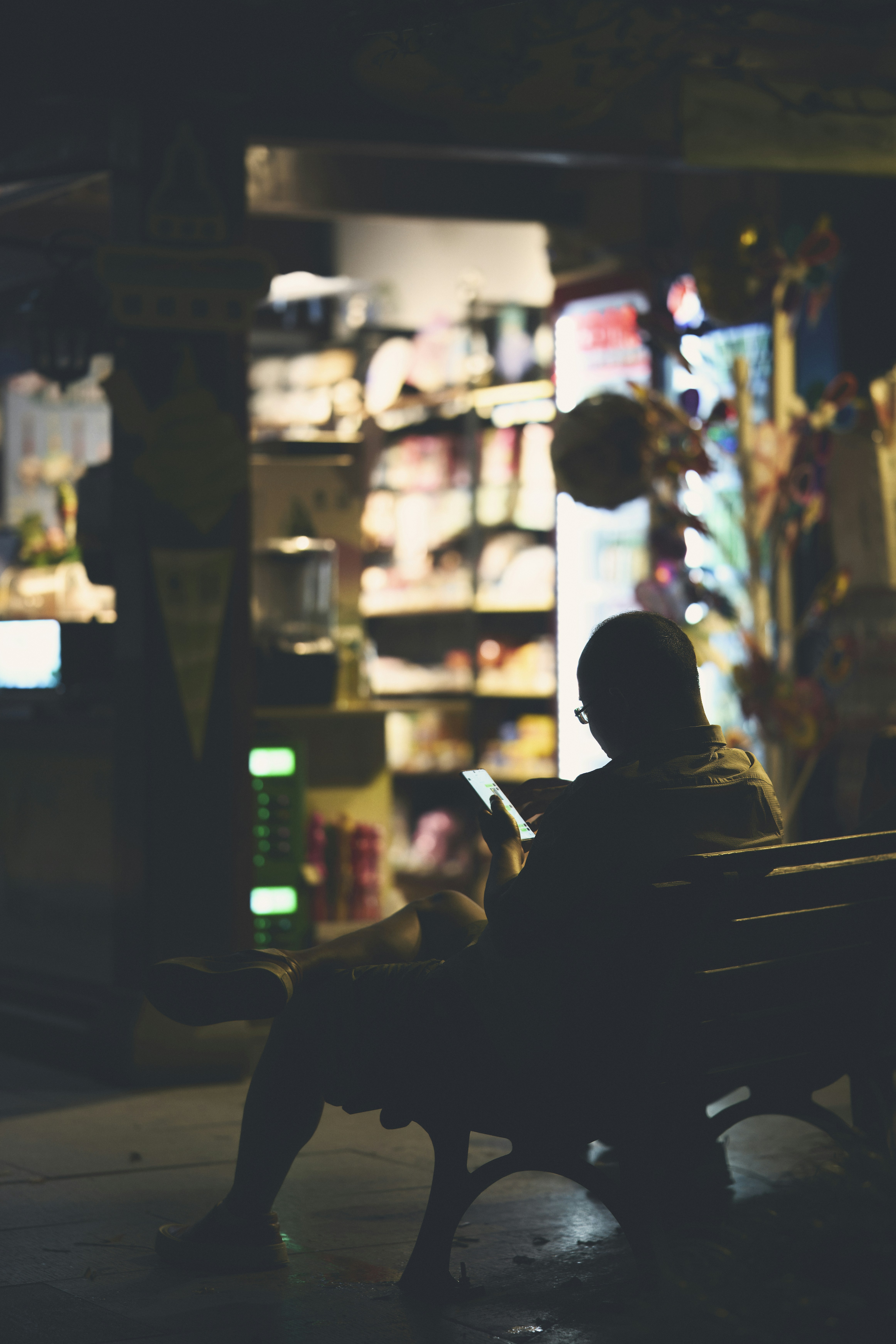 Person sitting on a bench, engrossed in their phone, illuminated by the soft glow of a nearby shop. The scene captures a moment of solitude amidst the vibrant backdrop.