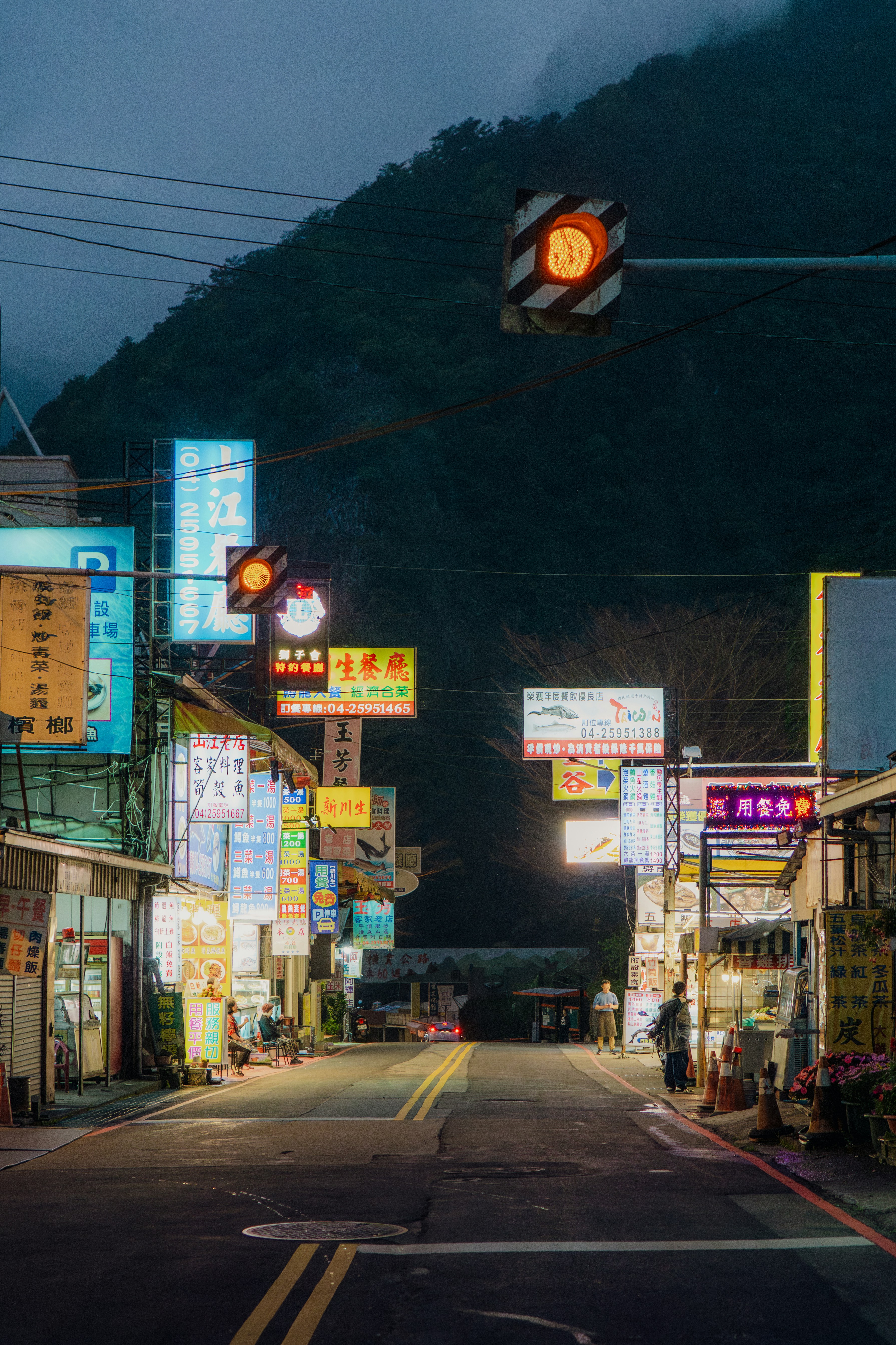 Vibrant street scene illuminated by neon signs and traffic lights, with a foggy mountain backdrop. The atmosphere captures the essence of nightlife in a bustling urban setting.