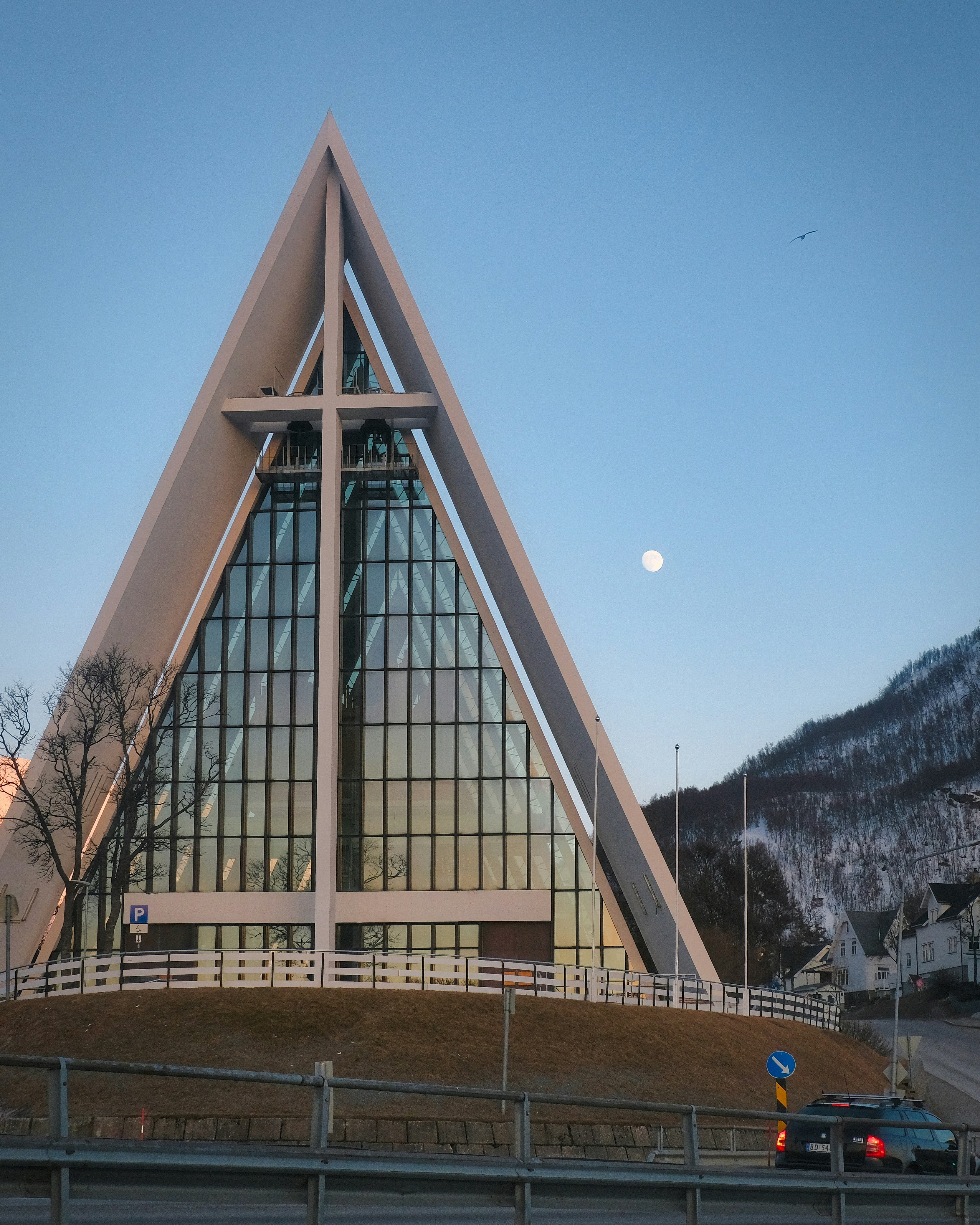 The Arctic Cathedral stands sharply against a clear sky at dusk, with the full moon rising beside its iconic triangular structure. This modernist church, officially known as Tromsdalen Church, is a well-known landmark in Northern Norway. | A modern church is visible with a moon in the sky.