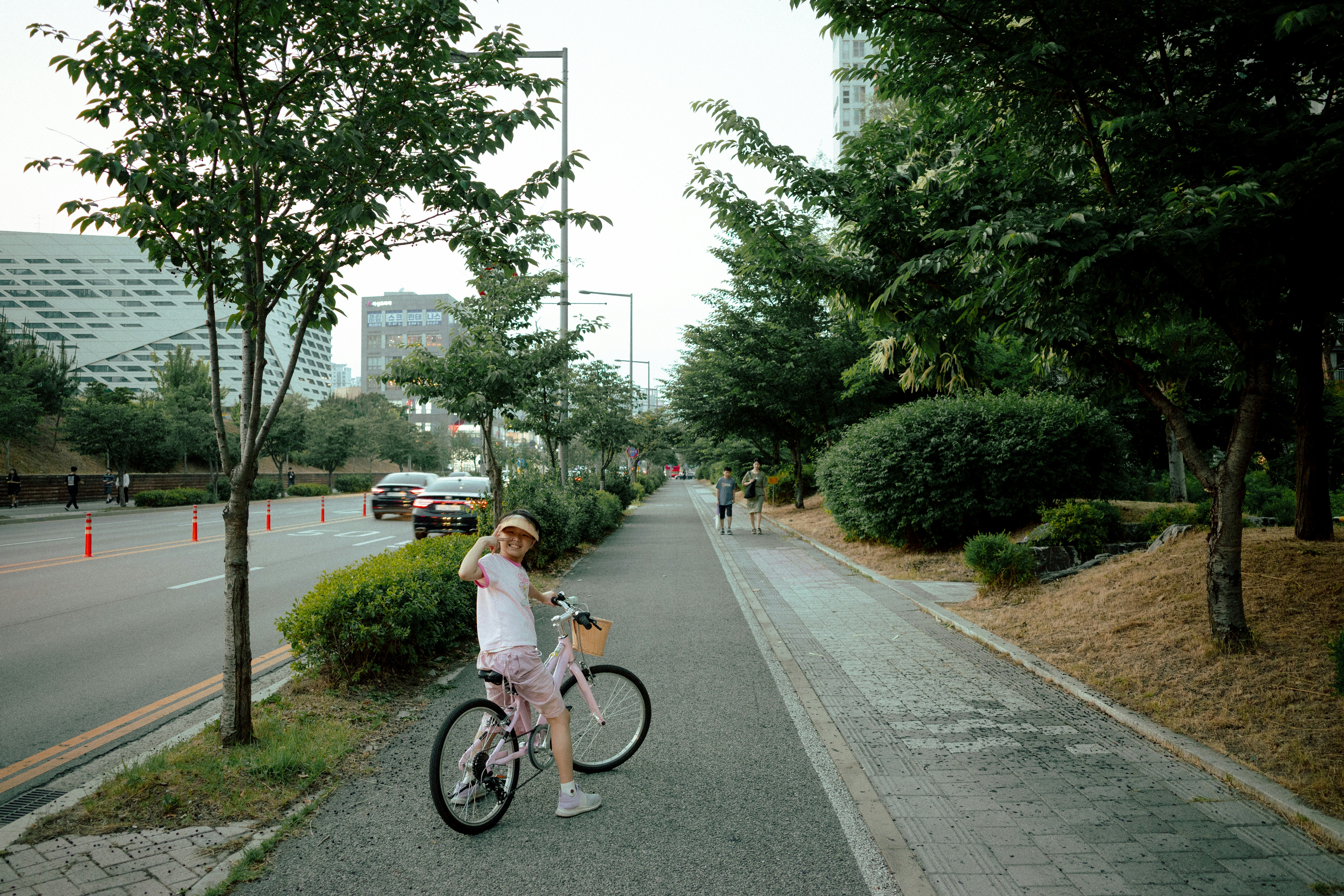 Child smiling and waving while sitting on a bicycle along a tree-lined path in an urban setting.