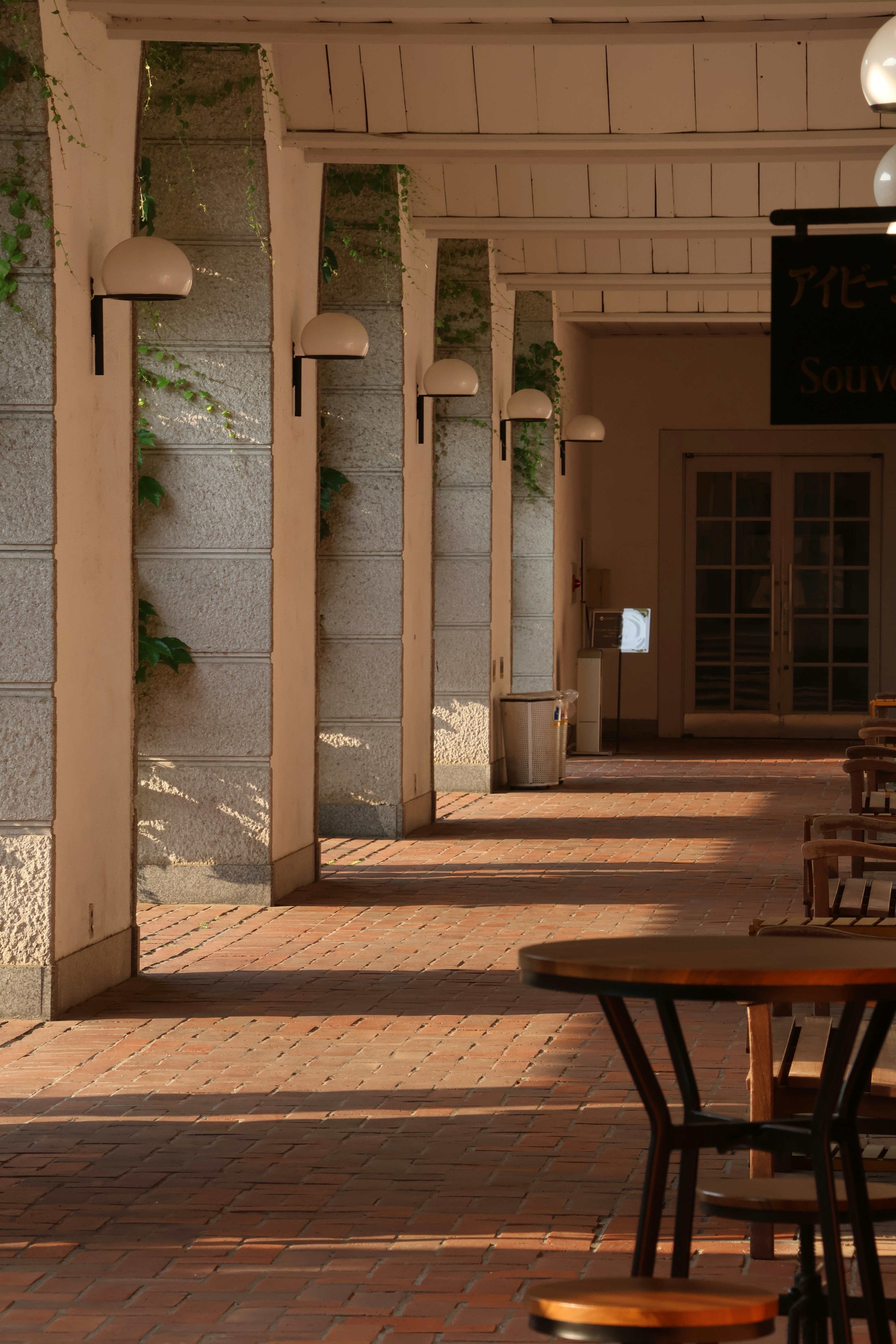 A serene arcade lined with columns and soft lighting, showcasing a quiet space with wooden tables and creeping greenery. The atmosphere invites calm reflection.