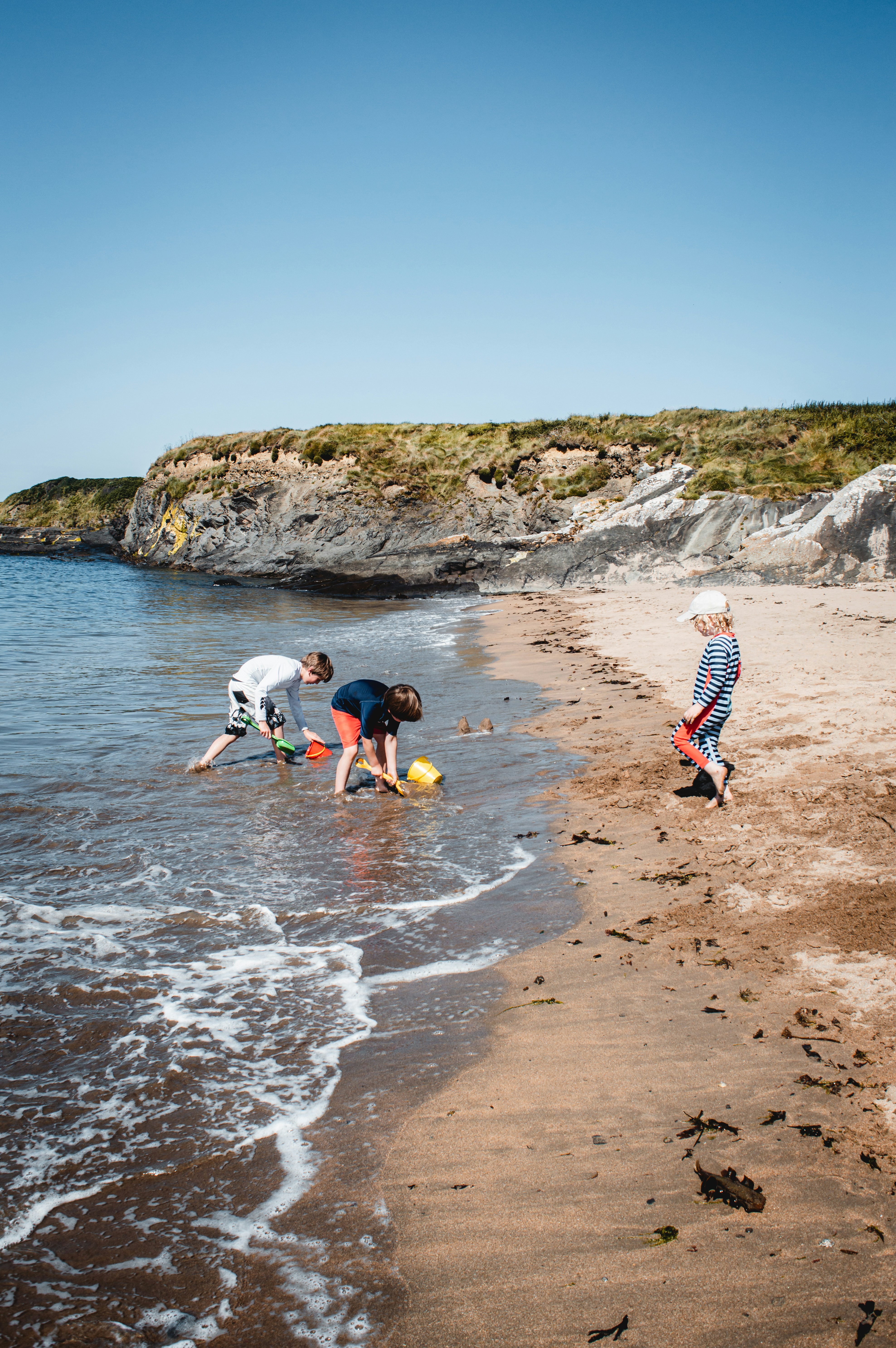 Three children playing on a sandy beach, engaging with the water and sand, while a rocky coastline looms in the background.