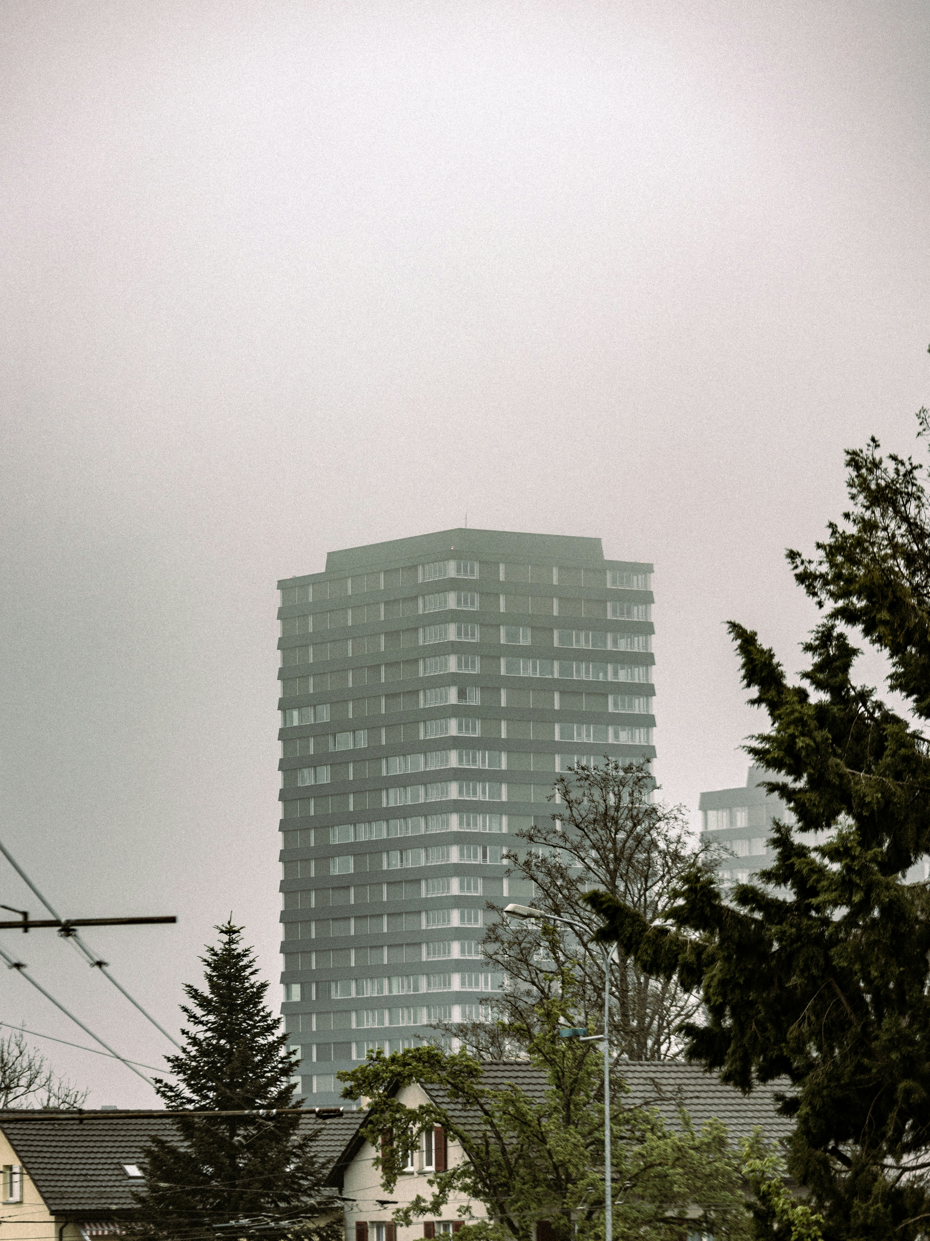 A modern high-rise building looms through a foggy atmosphere, surrounded by trees and rooftops, evoking a sense of urban mystery.