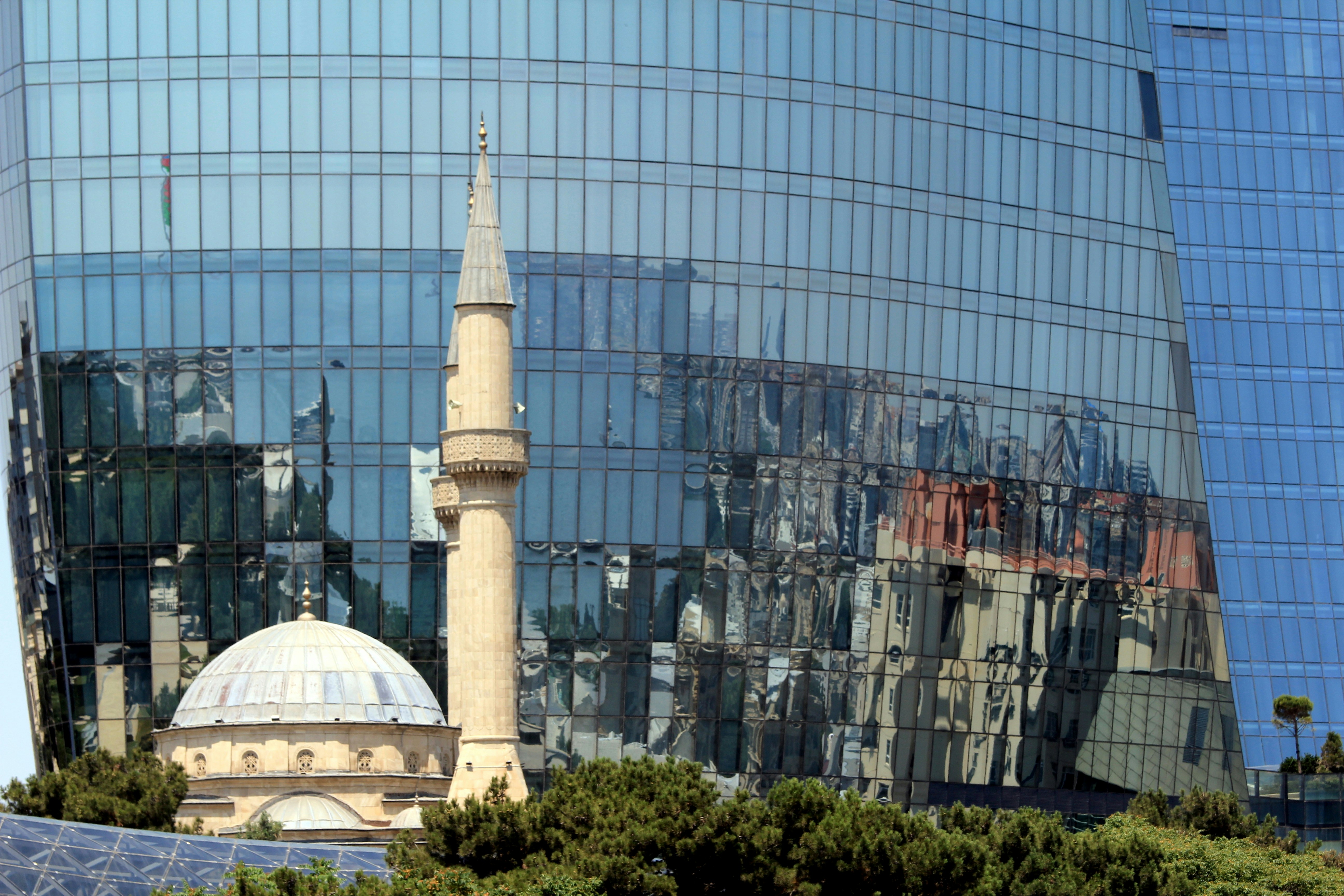 A historic mosque's minaret and dome reflected in the sleek glass facade of a contemporary building, illustrating the blend of architectural styles.