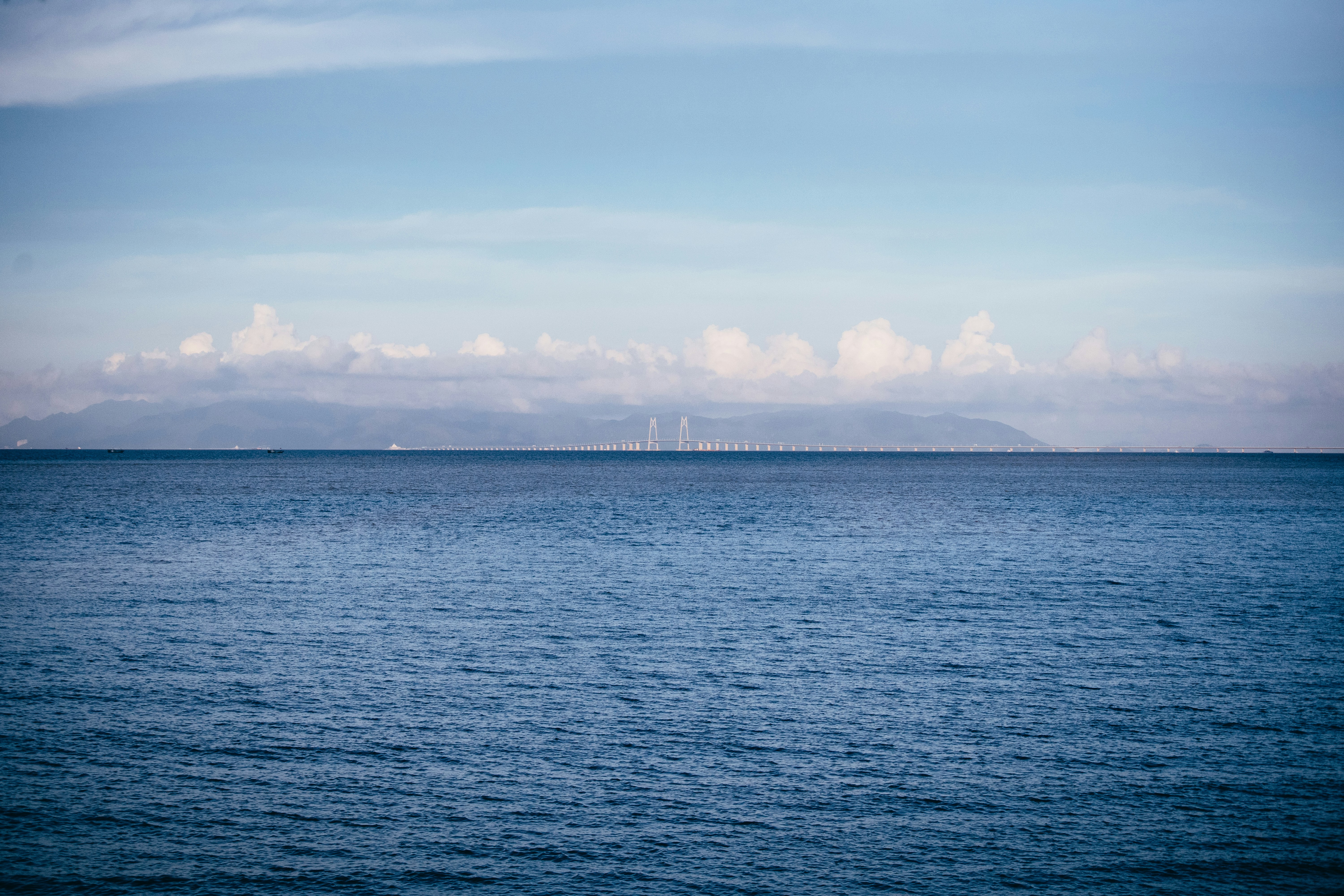 Distant bridge spans across a tranquil sea under a soft blue sky, framed by gentle clouds. The scene evokes a sense of calm and connection.