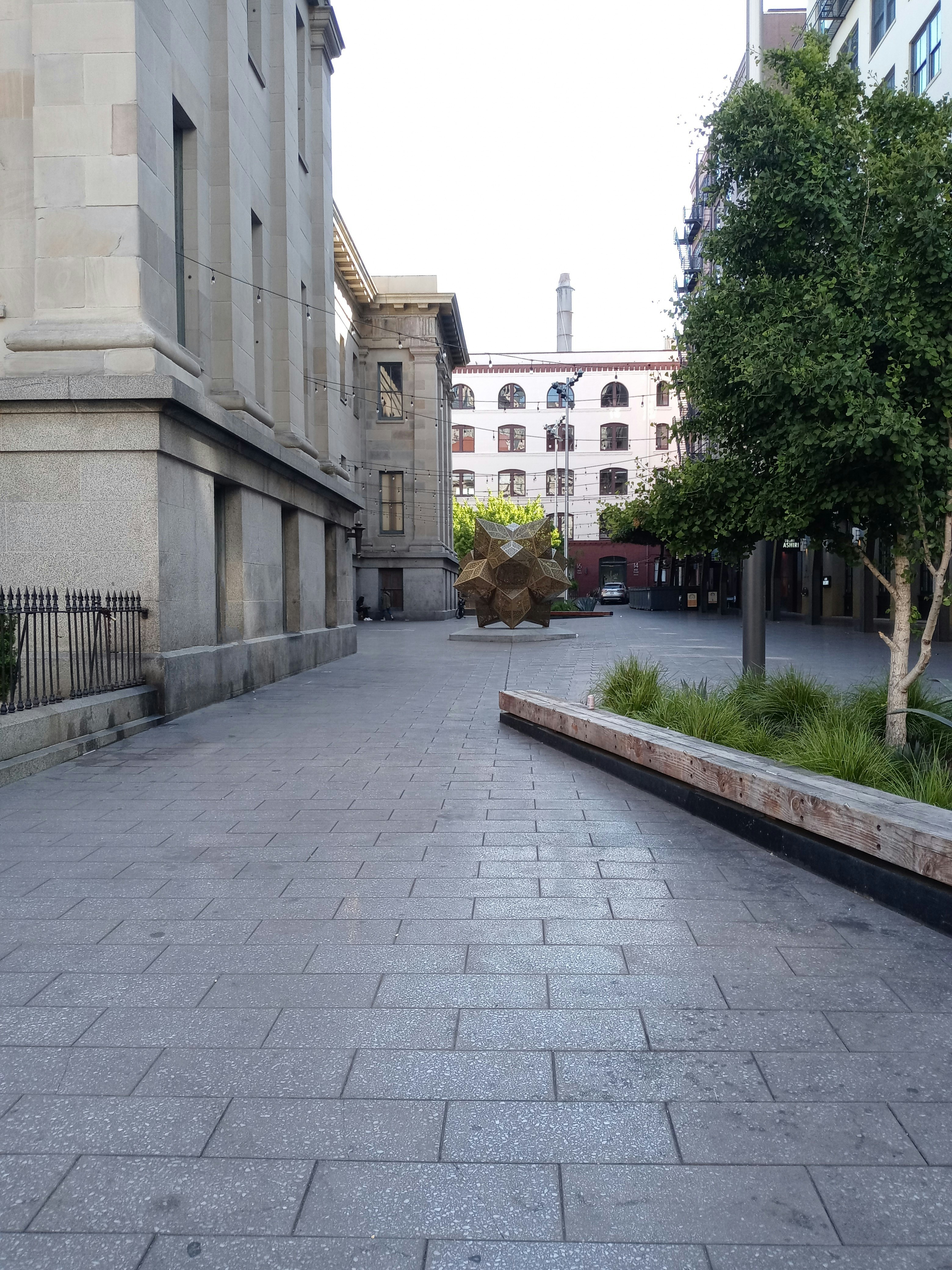 A modern urban pathway featuring a geometric sculpture surrounded by greenery and historic architecture. The scene captures a blend of contemporary art and classic building design.