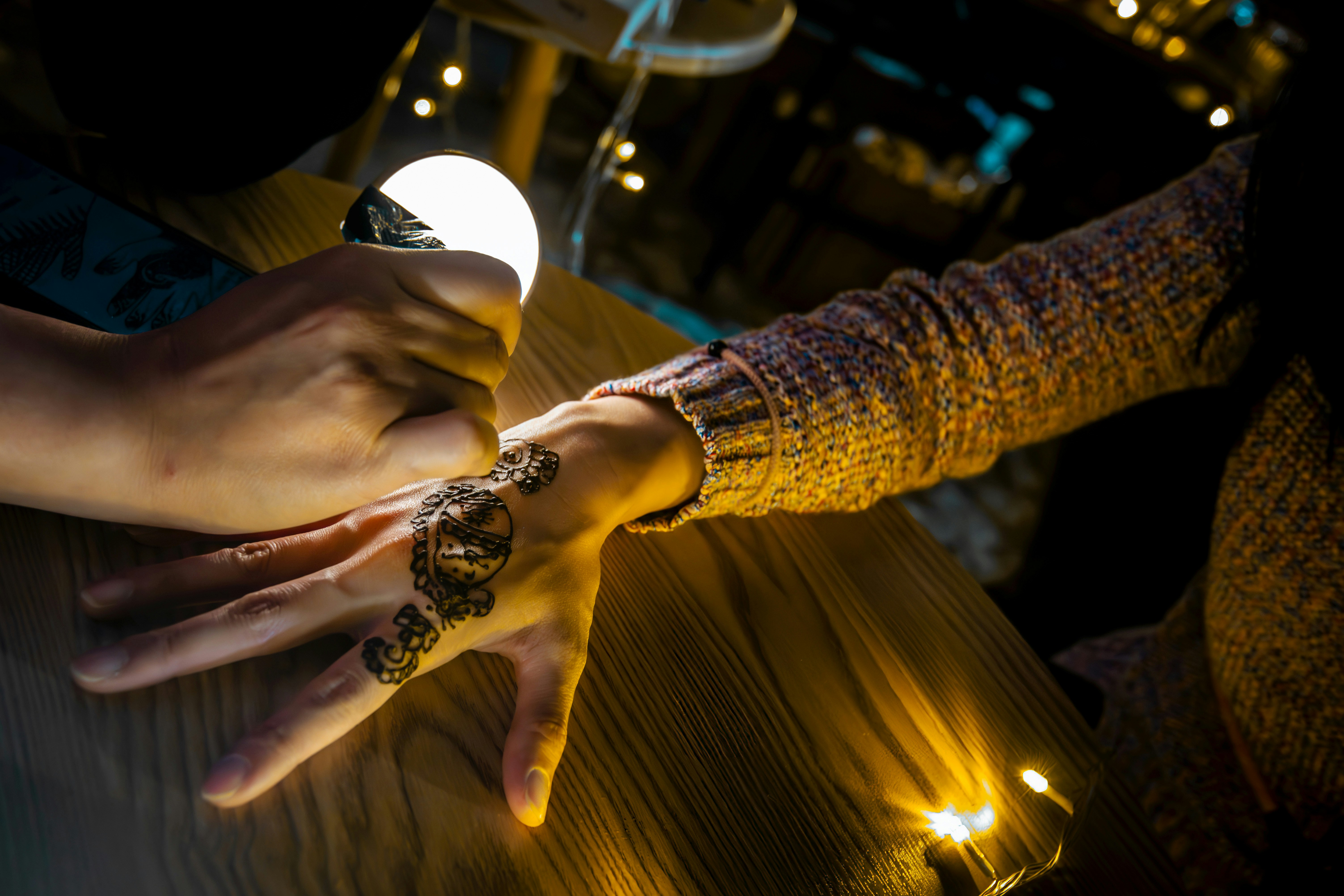 A hand adorned with detailed henna designs while being carefully painted, illuminated by a warm light source in a cozy setting.