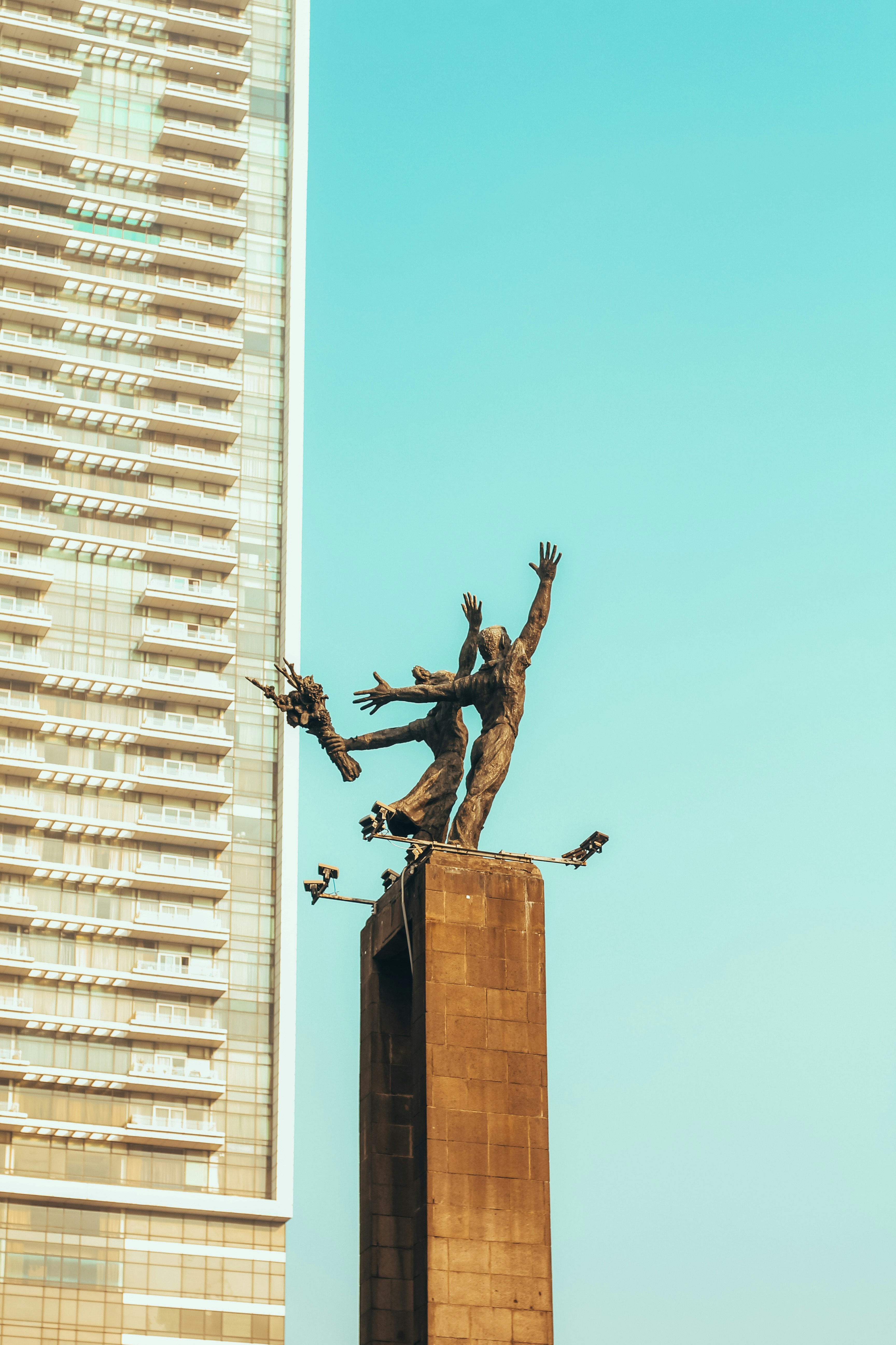 Bronze sculpture depicting a figure reaching out with arms extended, set against a contemporary building backdrop.