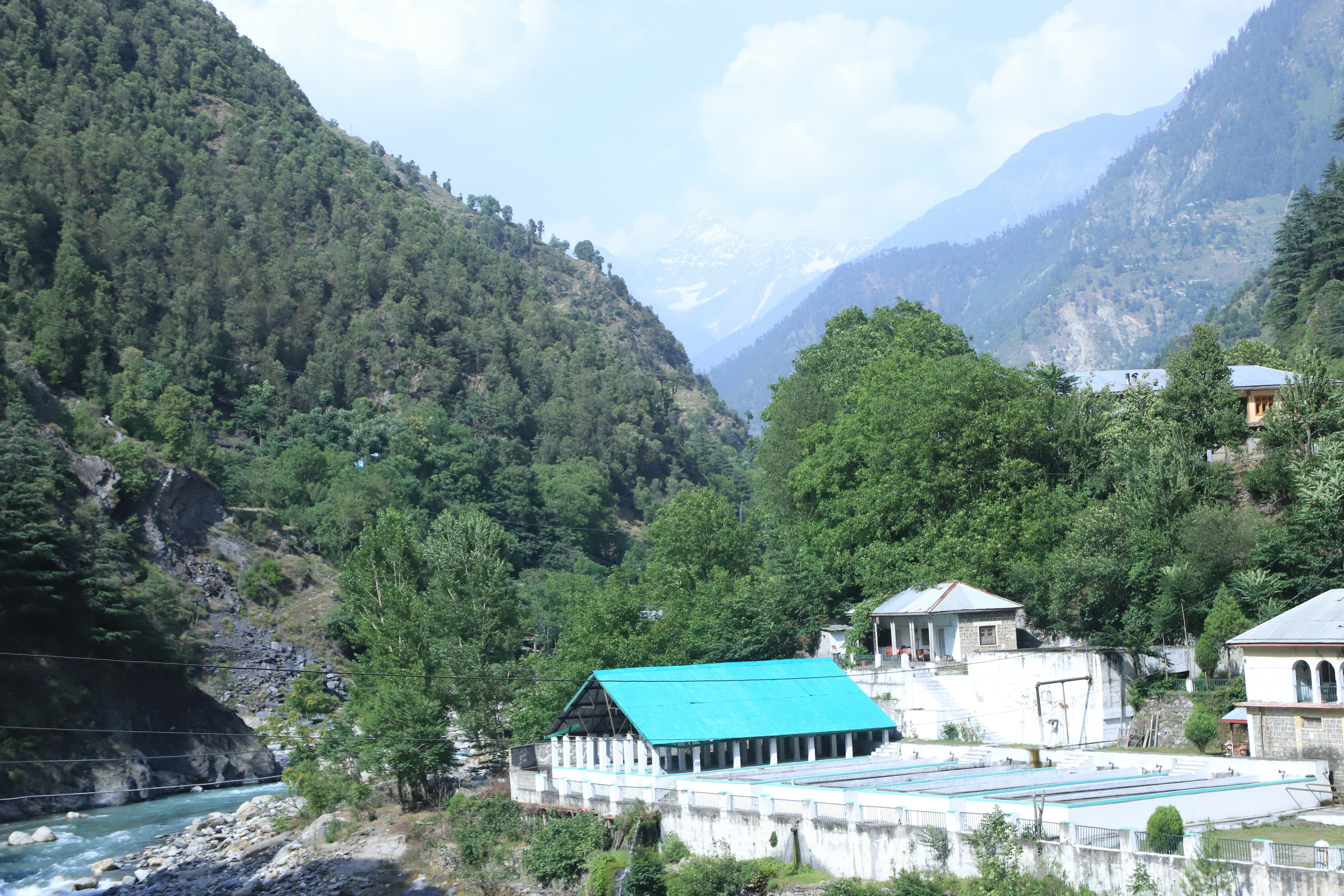 Lush green mountains frame a tranquil riverside scene with a quaint building featuring a blue roof. Snow-capped peaks peek through the clouds in the distance.