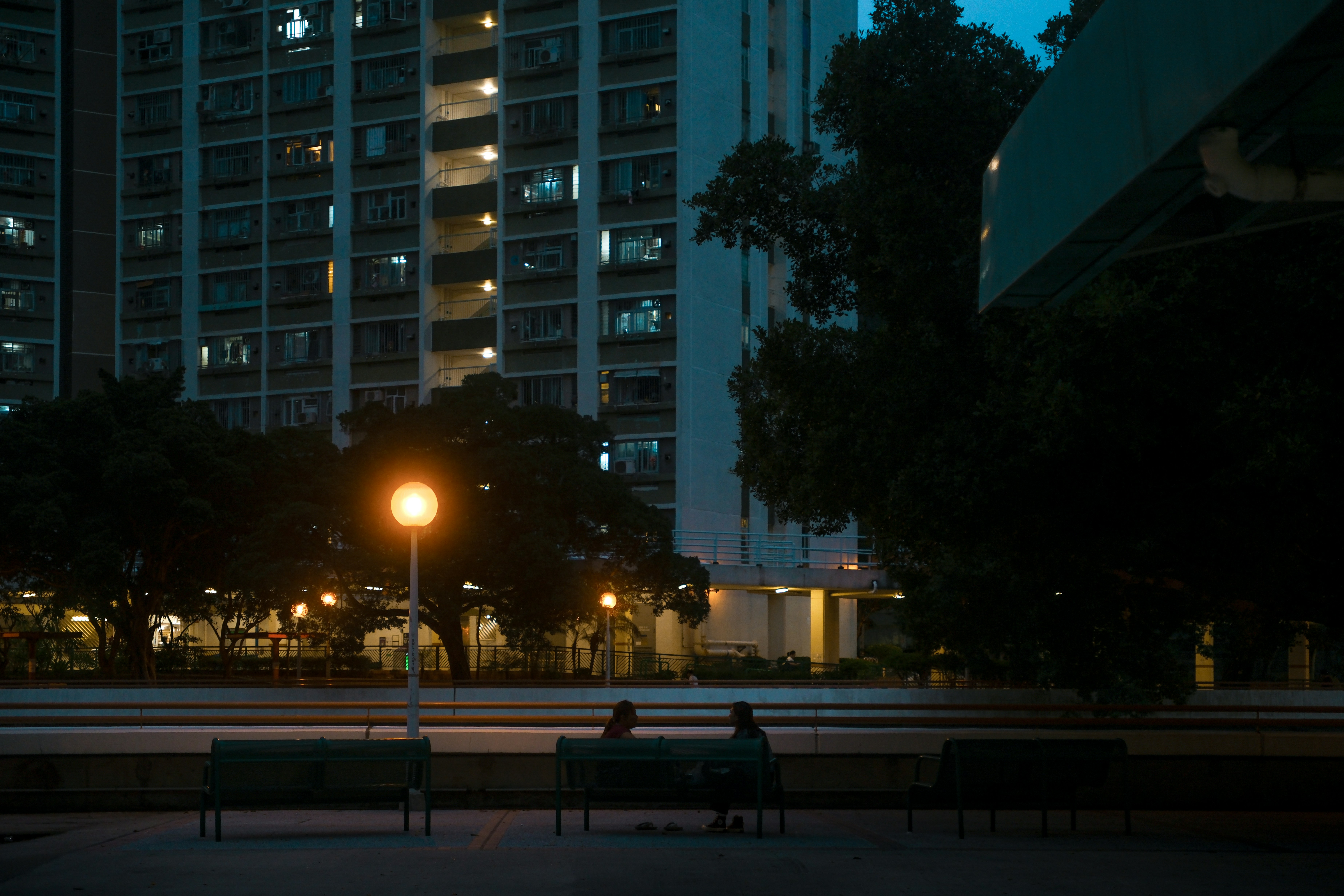Two figures seated on benches beneath a glowing streetlamp in an urban setting, surrounded by dimly lit buildings and trees.