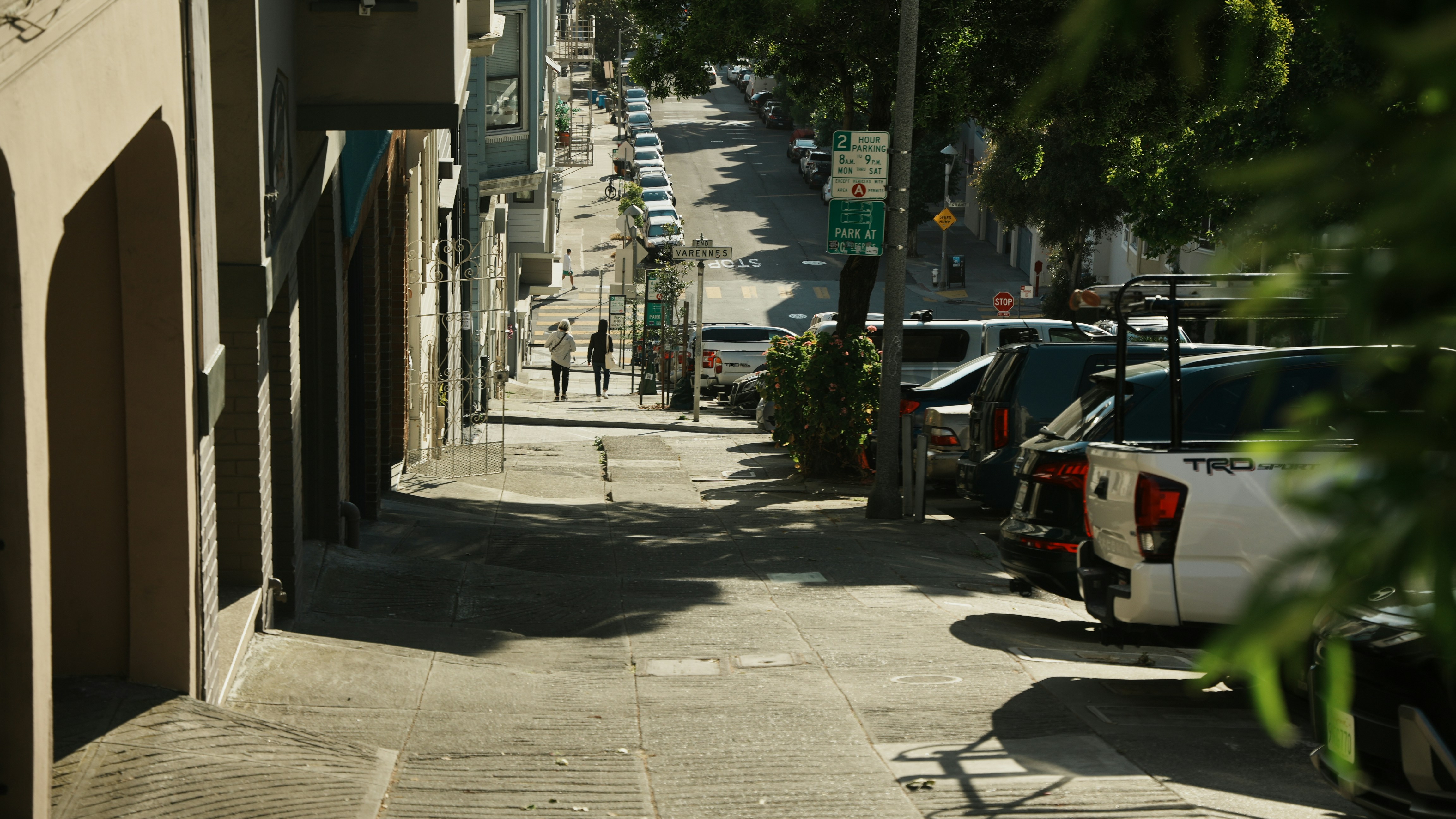 Two pedestrians walking down a sunlit, sloped street lined with parked cars and greenery.