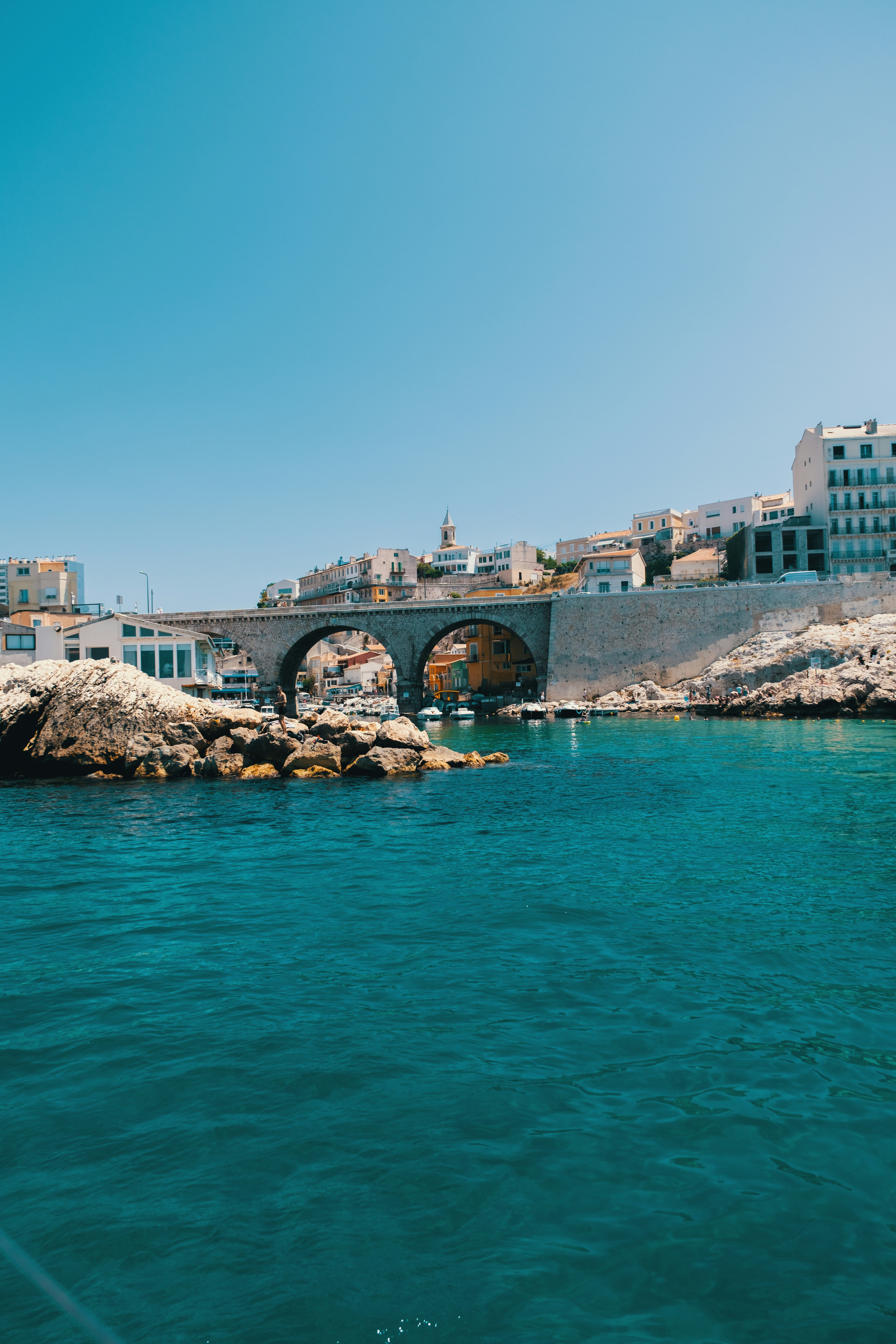 Historic stone bridge arching over tranquil waters with colorful buildings lining the shore. The scene captures a peaceful coastal town atmosphere.