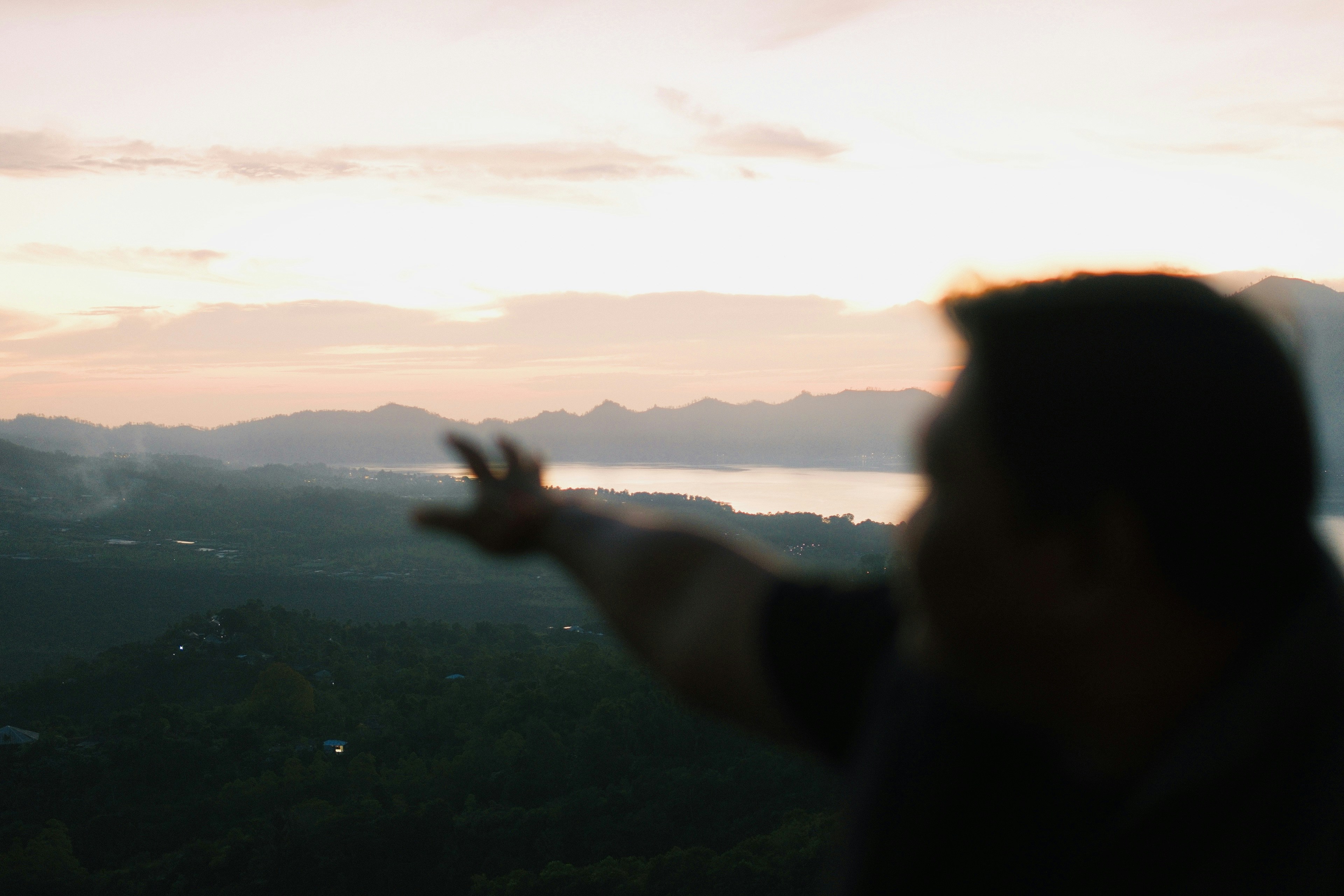 Silhouetted figure gesturing towards a hazy landscape at sunset, blending nature's beauty with human expression.
