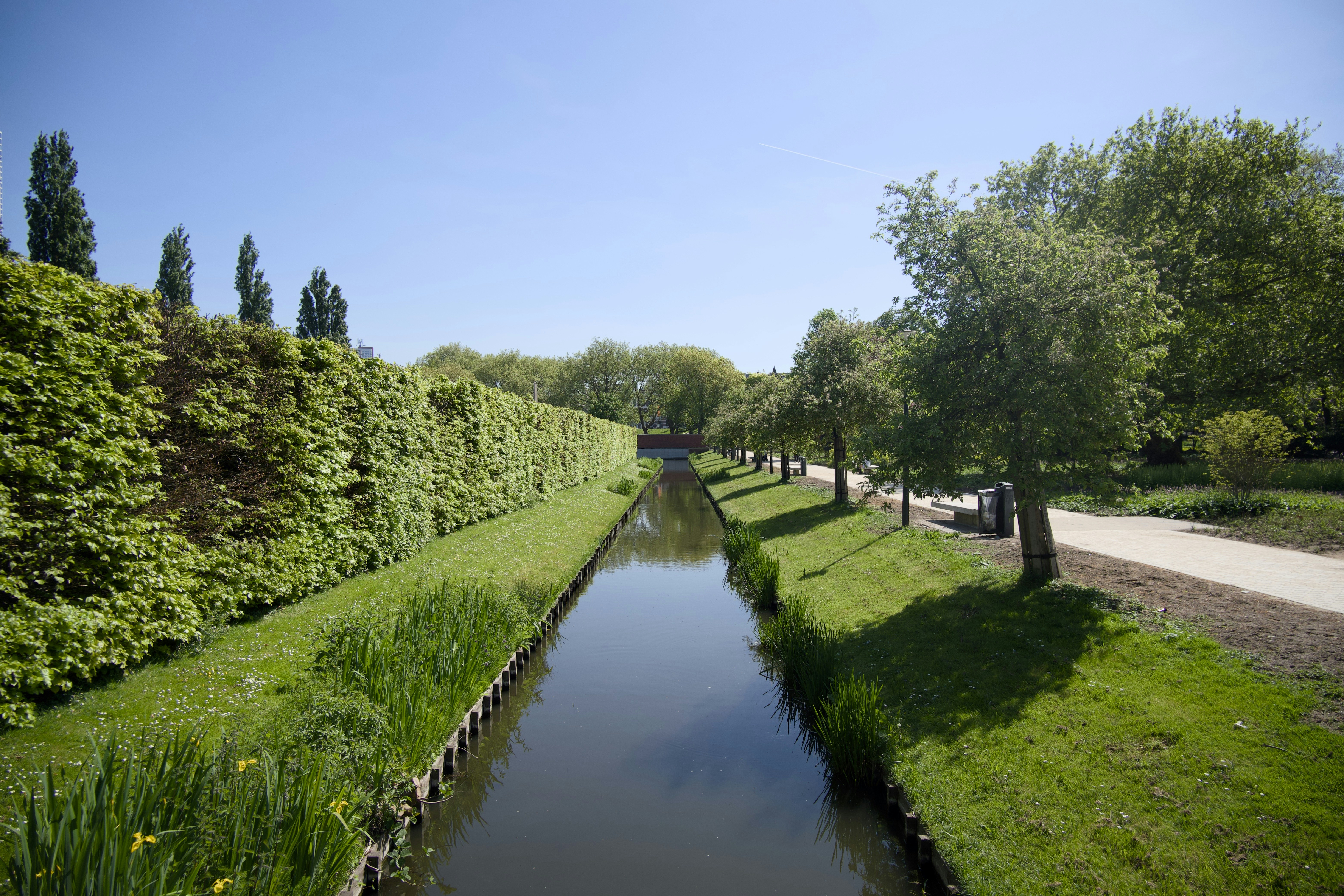 Lush greenery lines a tranquil canal, reflecting the clear blue sky above. A peaceful park path runs alongside, inviting leisurely strolls.