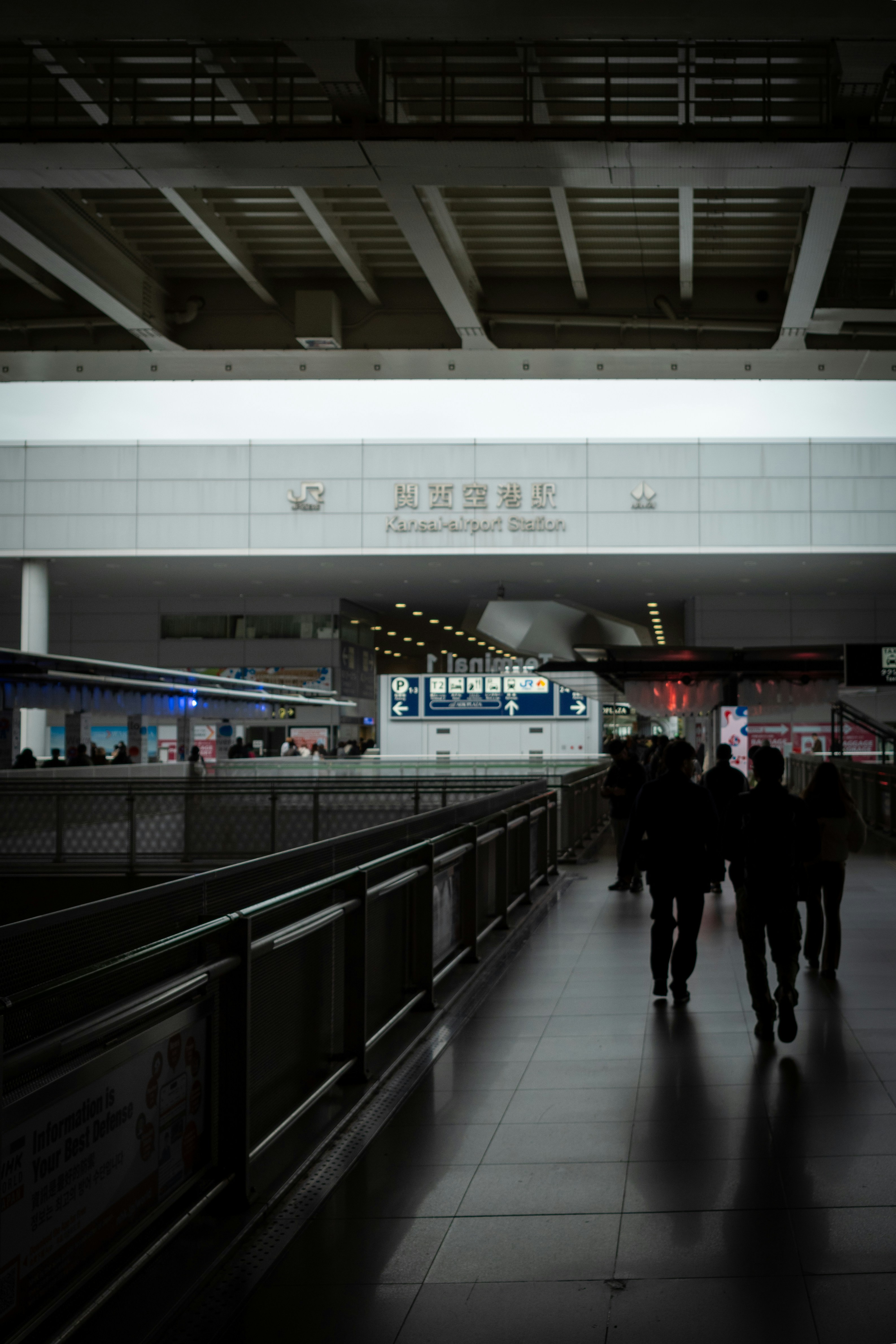 Silhouetted figures walking towards the entrance of an airport station, with illuminated signage and a contemporary architectural backdrop.