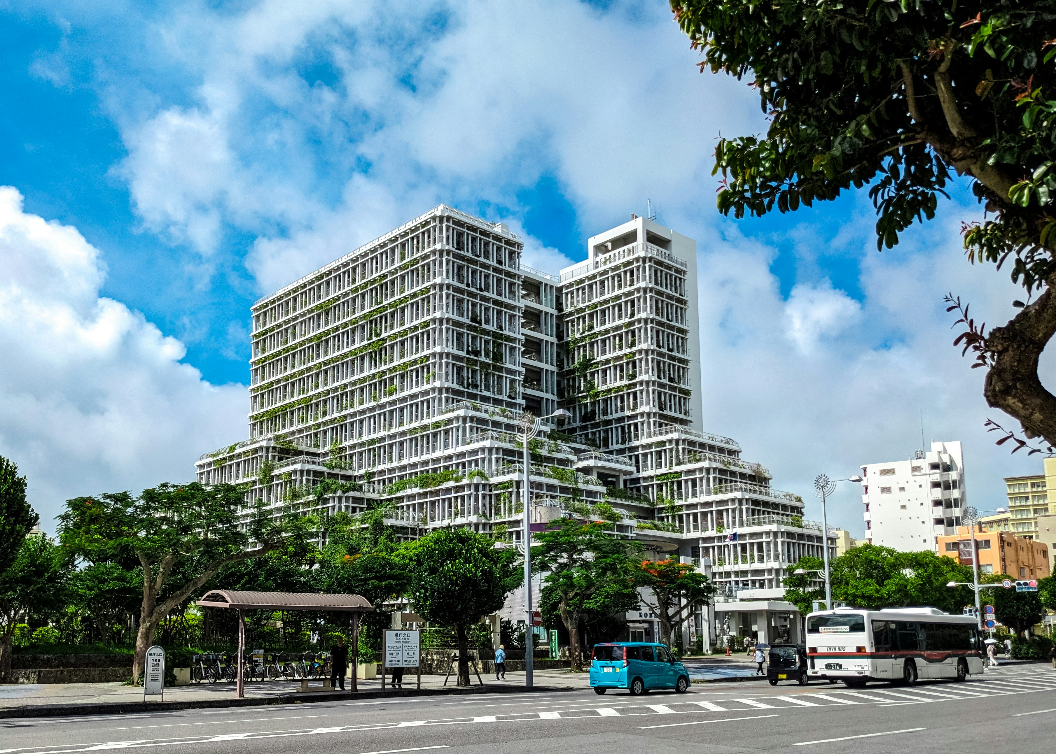 Compact cityscape featuring a terraced modernist building with greenery, street activity, and vehicles in front; a logo-like tree canopy frames the scene.