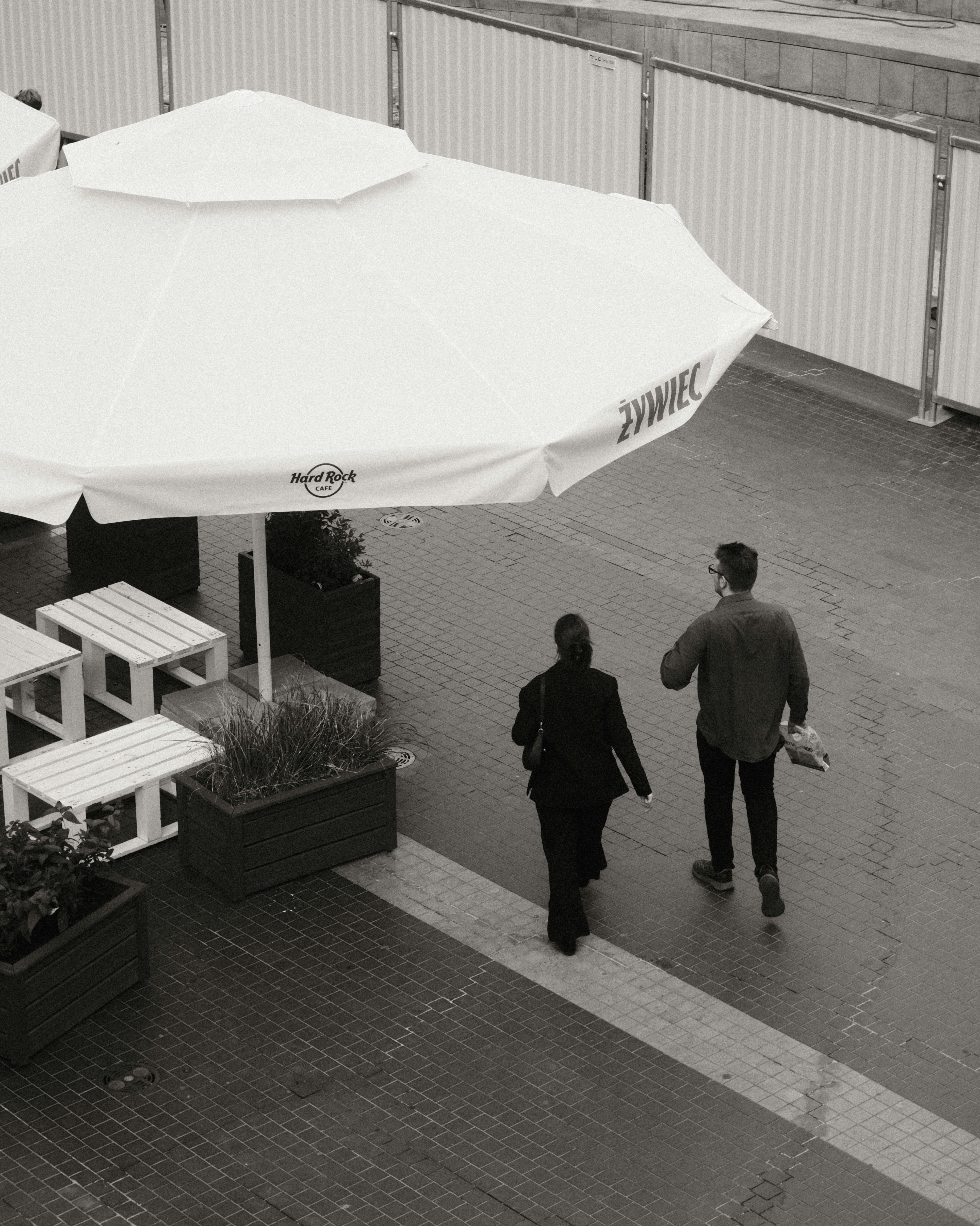 Stride in Shadow: Two Figures Under a Café Umbrella