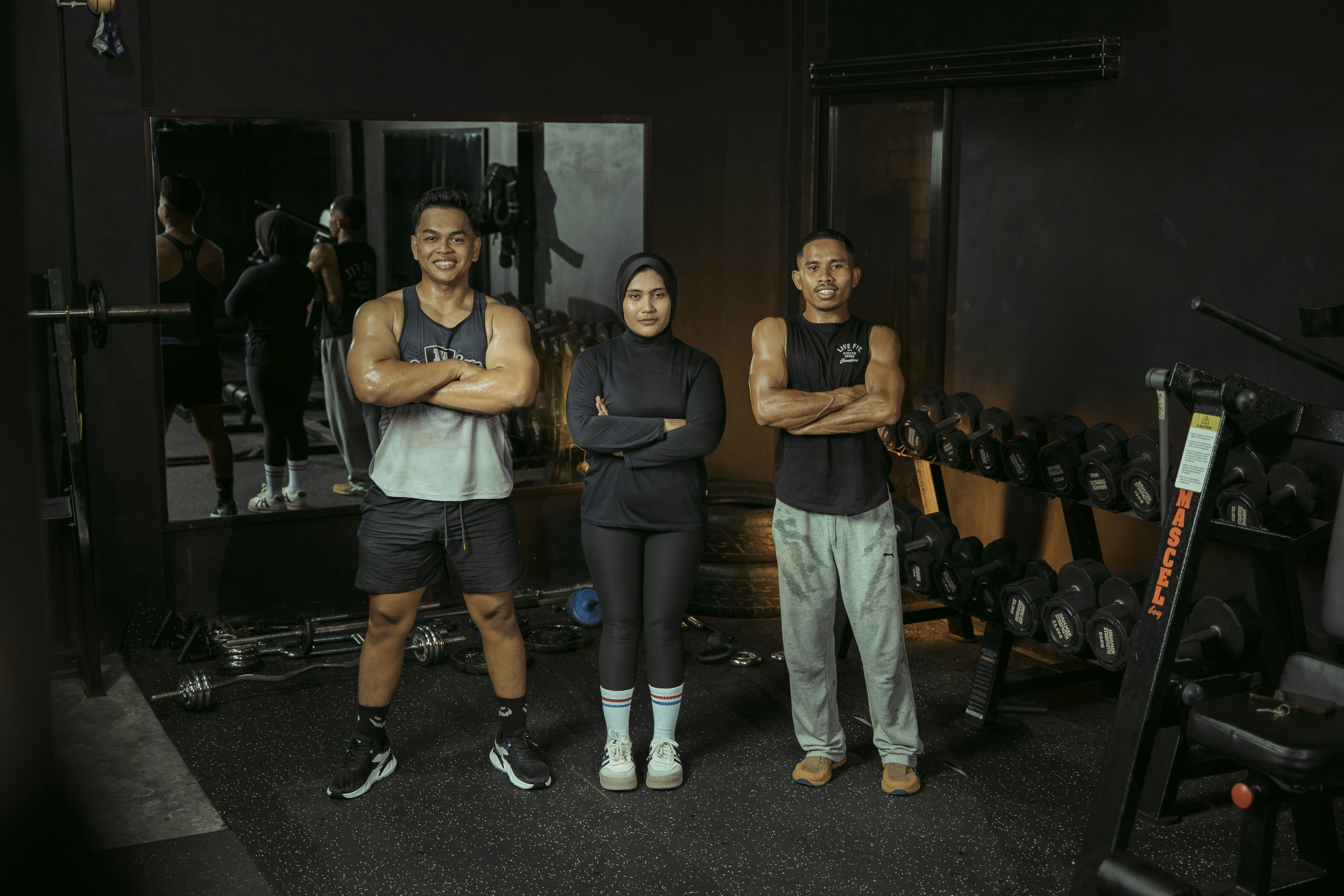Three fitness enthusiasts posing confidently in a gym, showcasing their dedication and camaraderie. The backdrop features weights and mirrors, emphasizing their commitment to health.