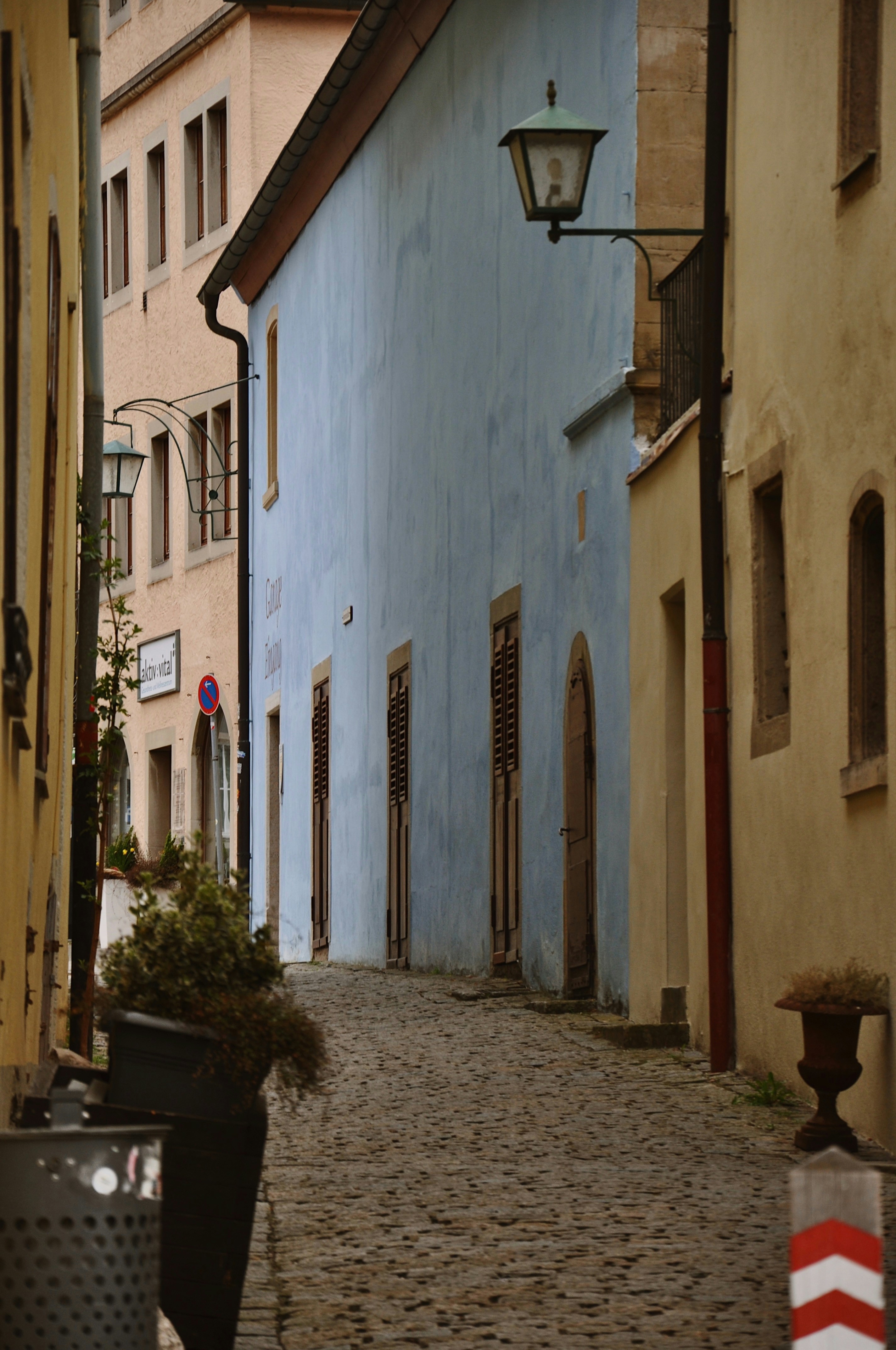 Narrow cobblestone alleyway flanked by pastel-colored buildings, with vintage street lamps adding charm. A potted plant adds a touch of greenery.