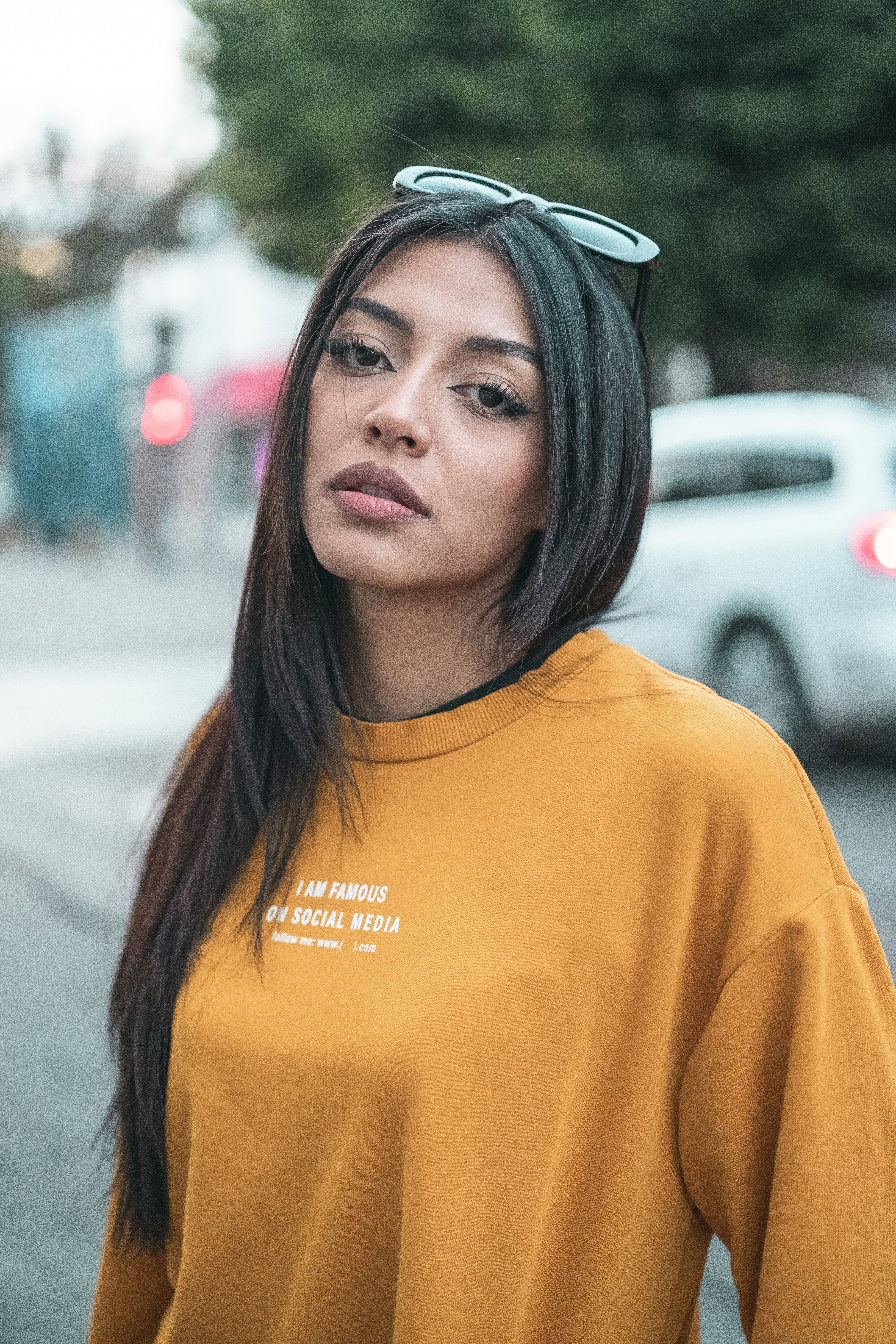 A young woman in a mustard sweatshirt gazes thoughtfully at the camera, with sunglasses perched on her head, set against a softly blurred urban backdrop.