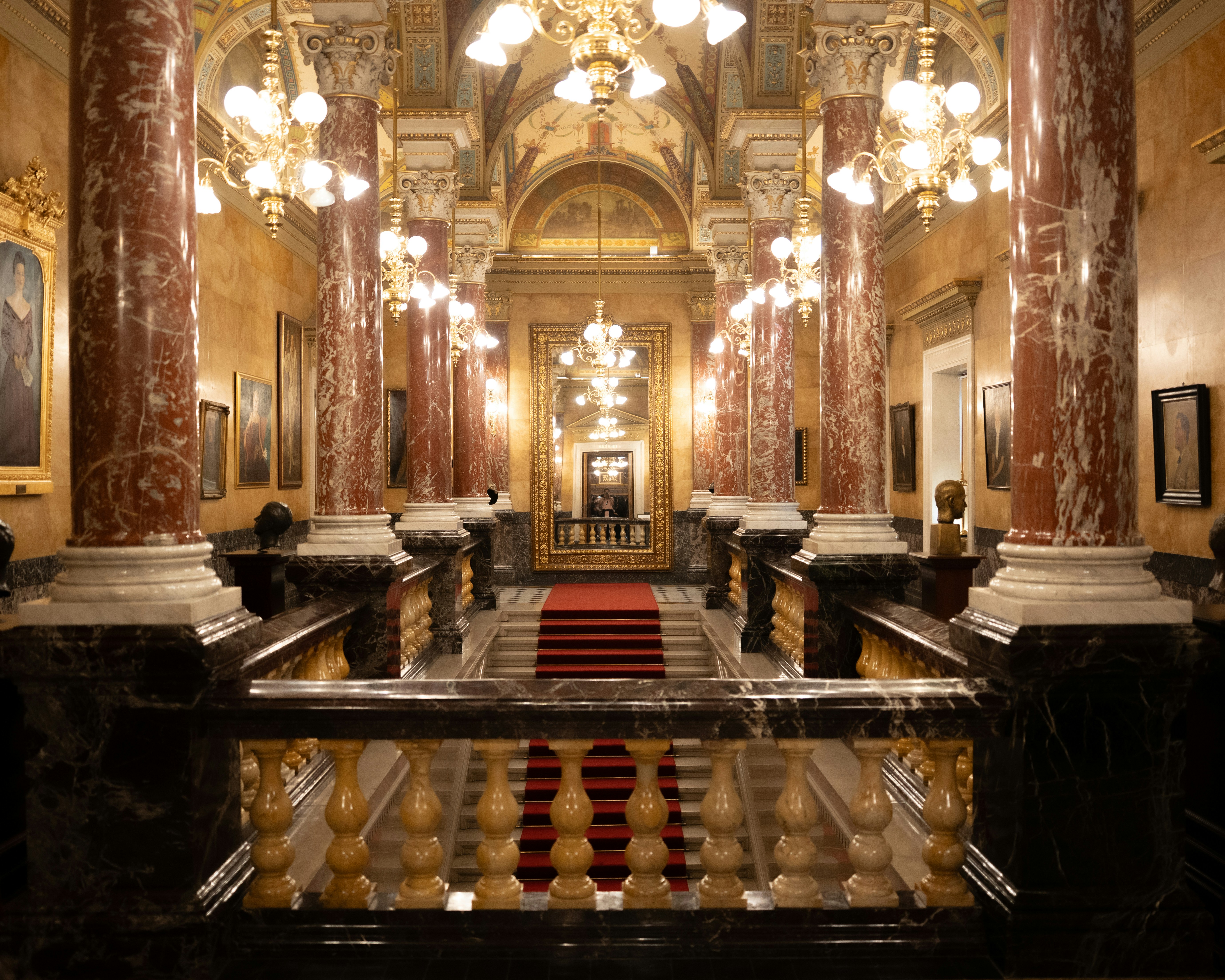 Luxurious interior of a grand hall featuring ornate columns, chandeliers, and a rich red carpet leading through the space.
