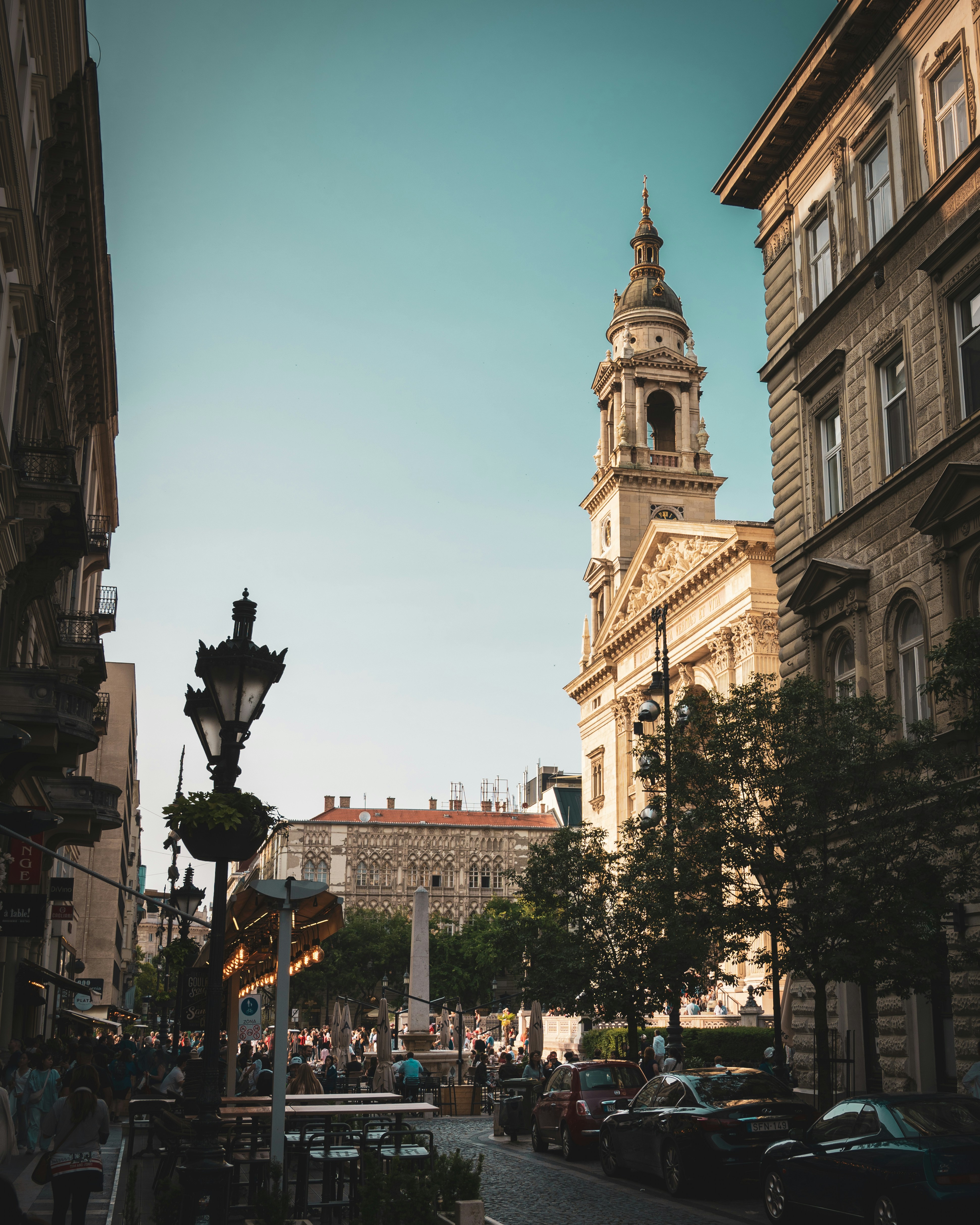 Historic building with a prominent clock tower rises above a bustling street filled with trees and pedestrians. Warm sunlight bathes the scene, highlighting the architectural details.