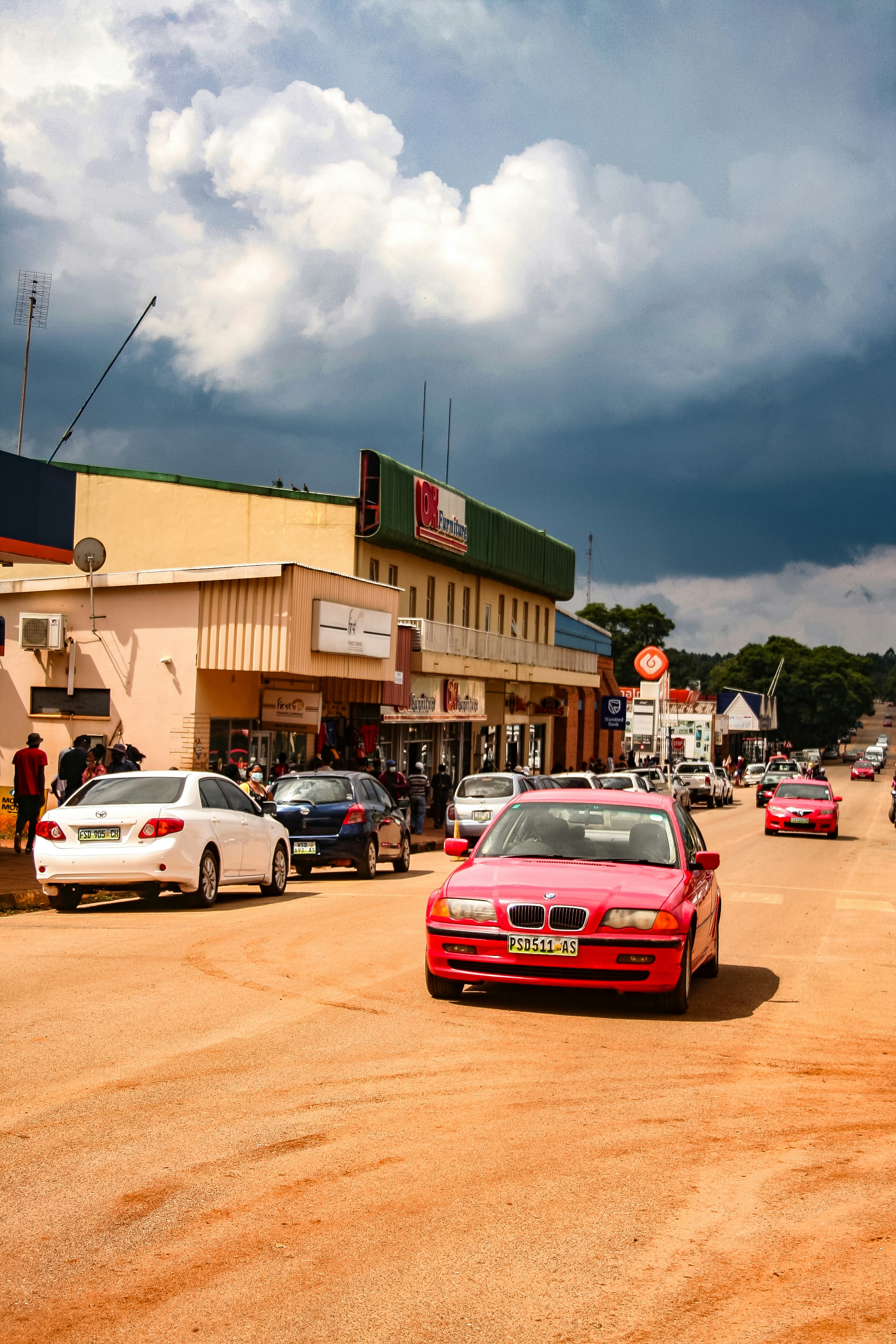 Red Sedan Leads a Dusty Town Street as Storm Clouds Gather