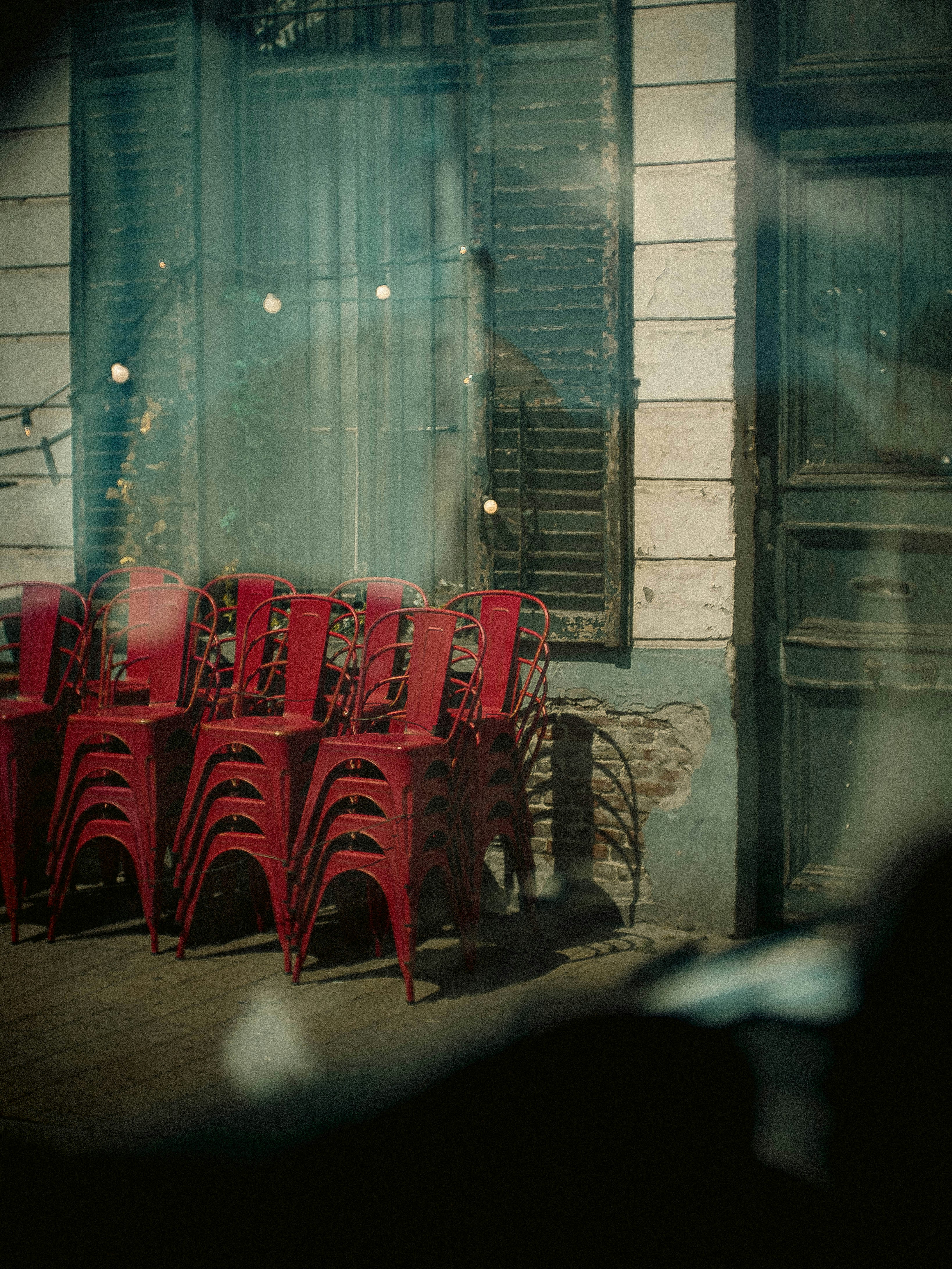 Stacked red chairs against a weathered wall, framed by a window with soft reflections. A glimpse of a rustic door complements the scene.