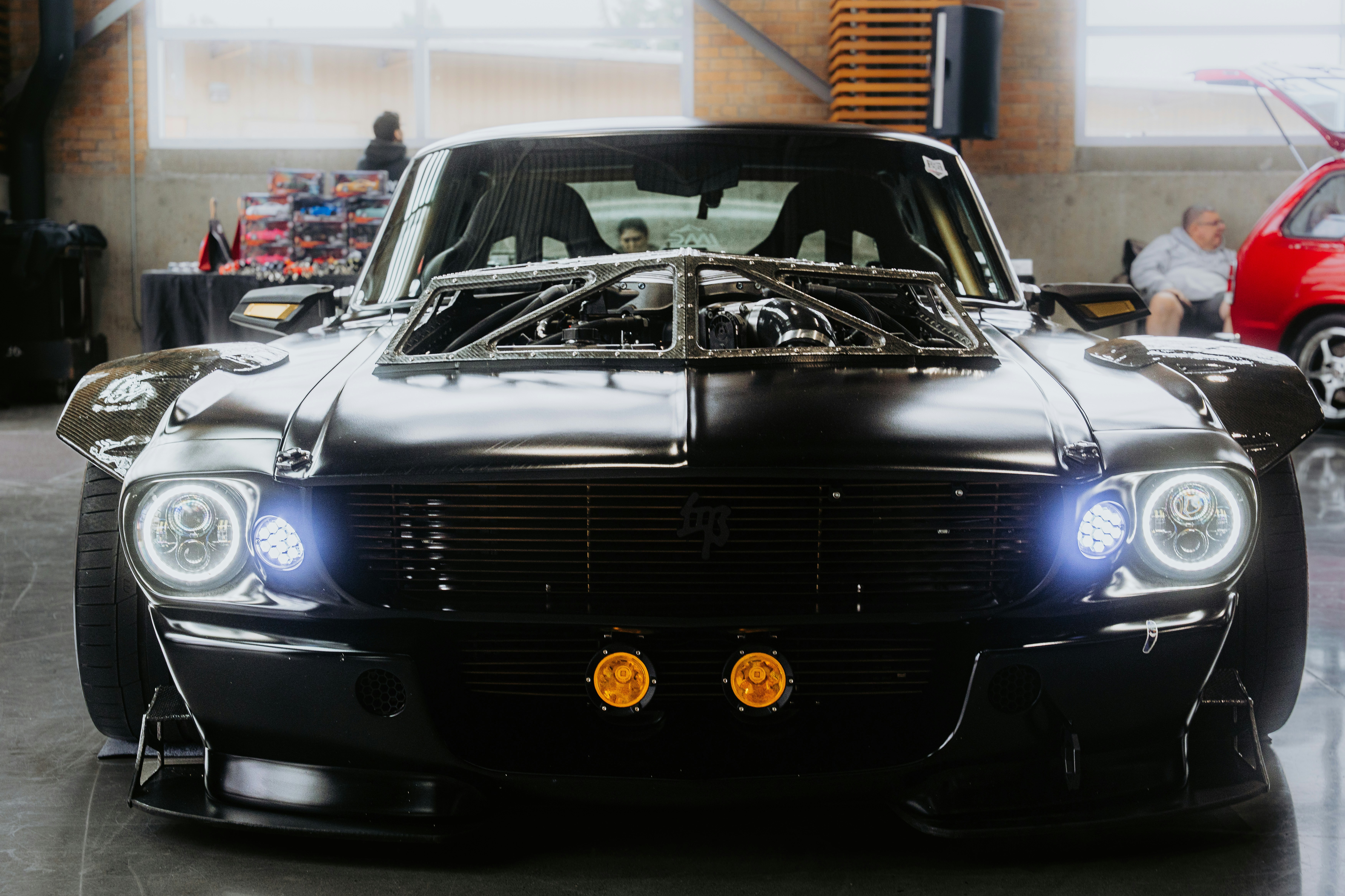 Modified black muscle car with a sleek design and prominent headlights, showcasing its powerful engine and unique styling. A vibrant car show backdrop enhances the automotive atmosphere.