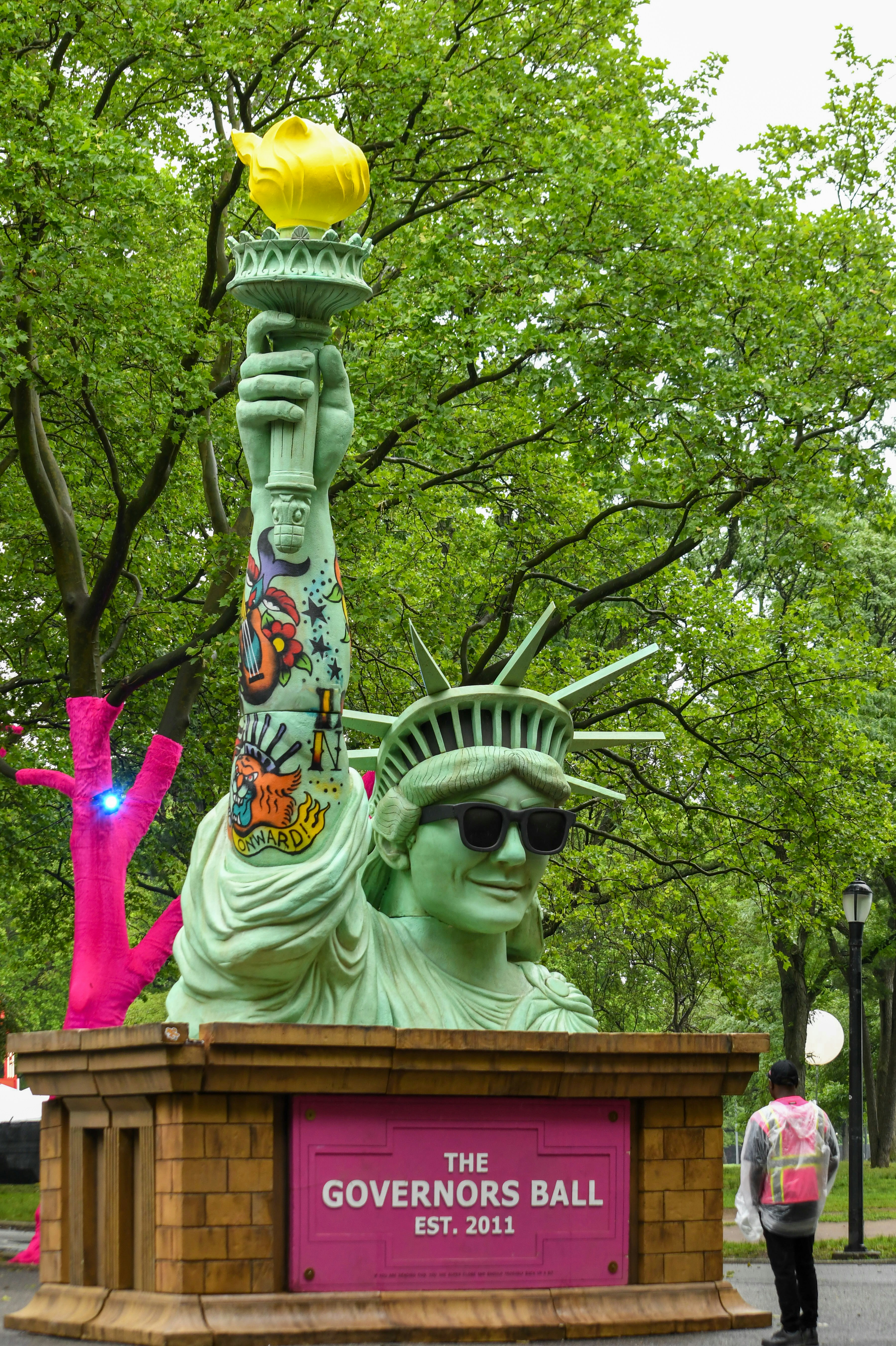 Statue resembling the Statue of Liberty adorned with colorful tattoos and sunglasses, set against a backdrop of vibrant greenery. The installation celebrates the Governors Ball festival.
