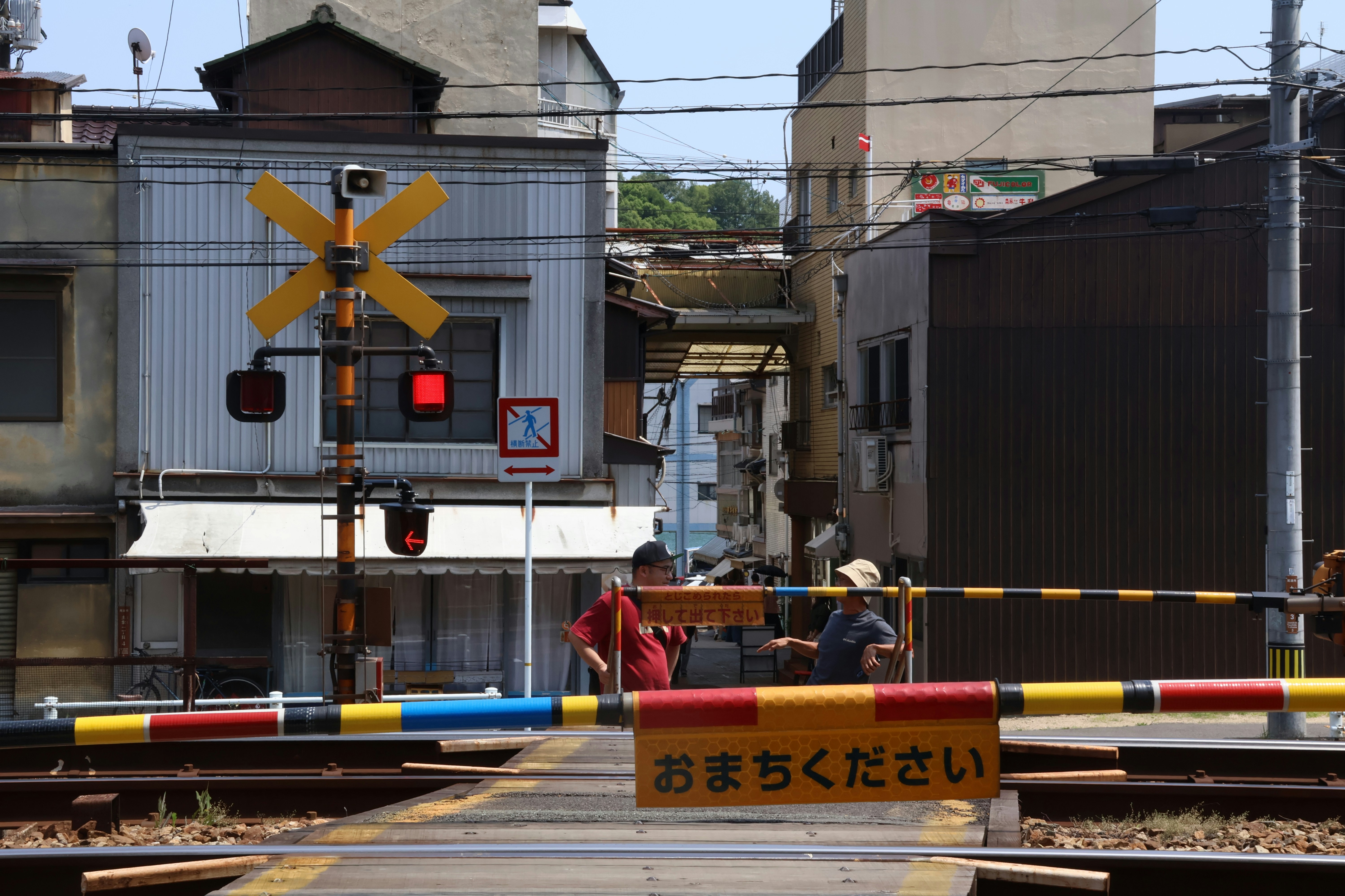 two men are talking aside the San-yo Line as they wait for the passing of an approaching train. | Train crossing with a warning light.