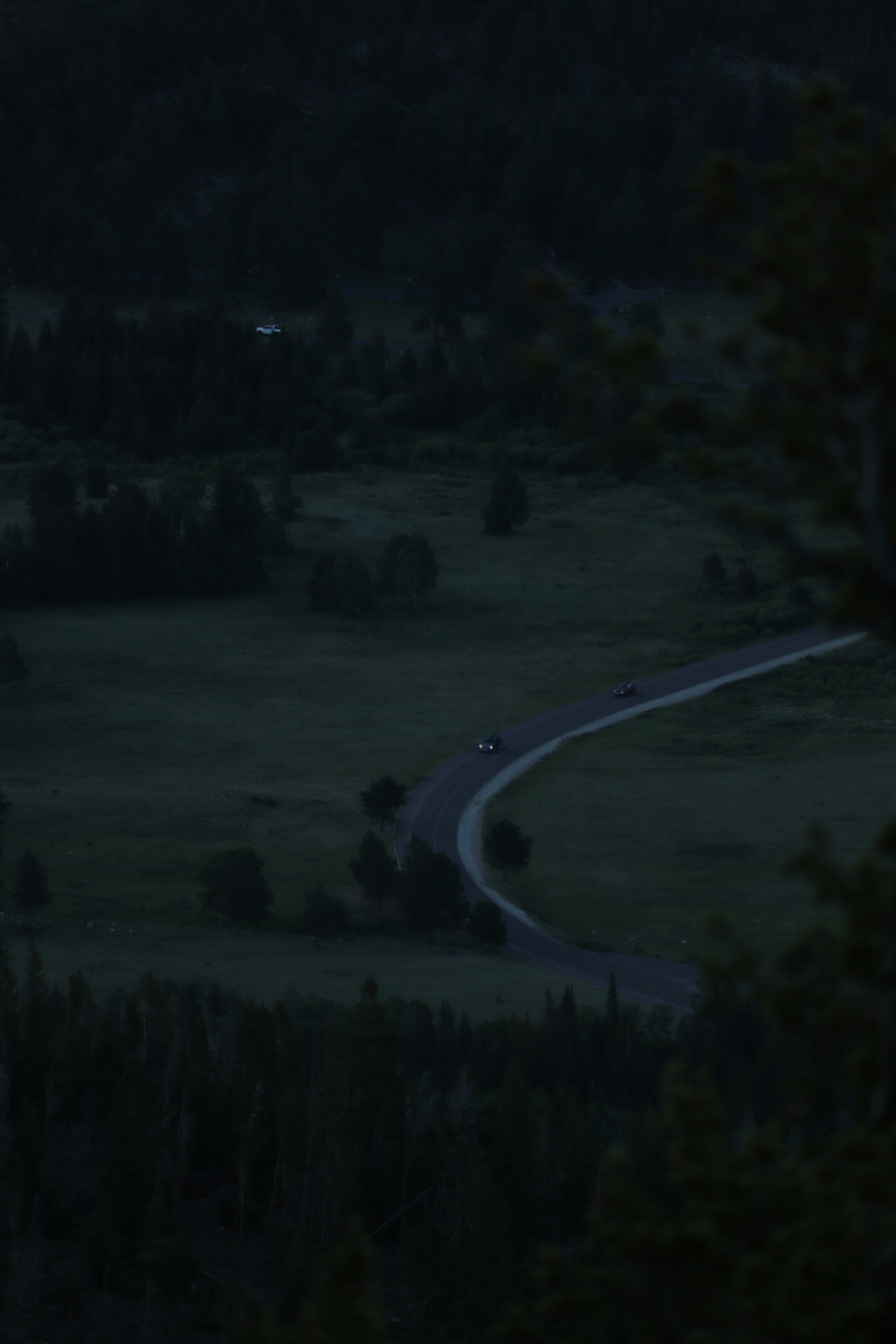 A winding road meanders through a serene valley, illuminated by distant vehicle headlights under a twilight sky.