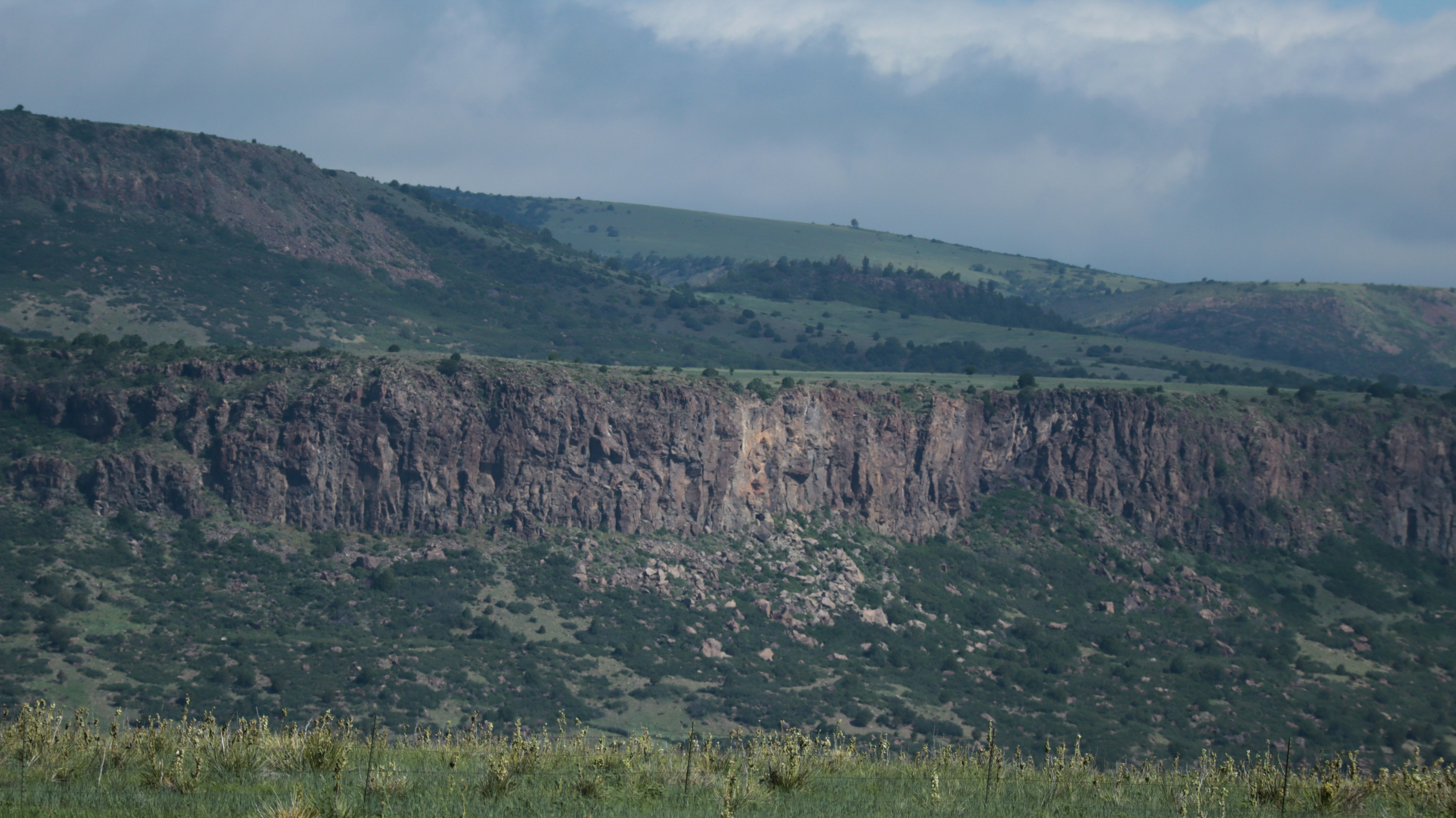 A rugged cliffside rises majestically against a backdrop of rolling hills and a cloudy sky, showcasing the beauty of natural landscapes.