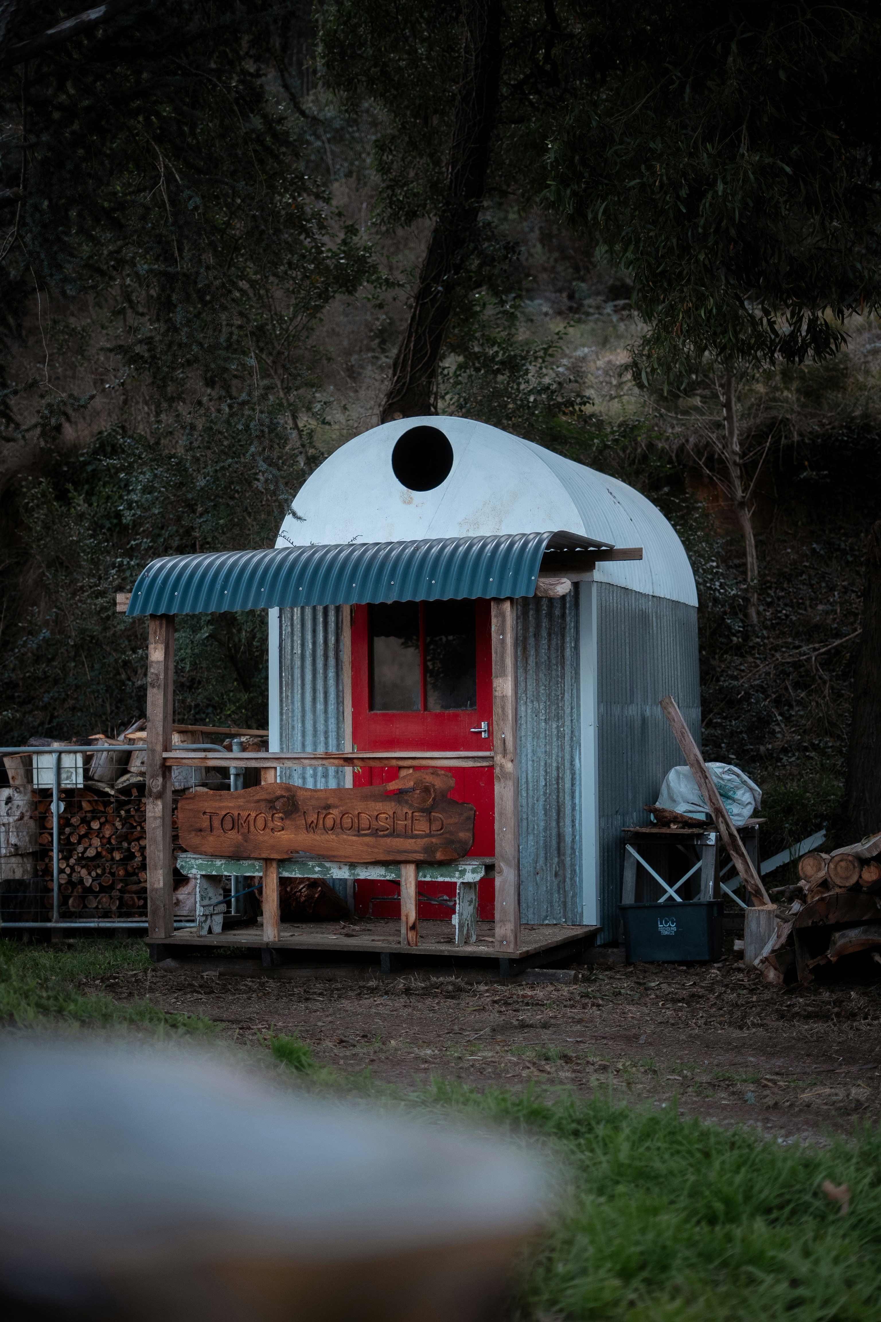 Quaint woodshed with a red door and corrugated metal roof nestled among logs in a serene natural setting.