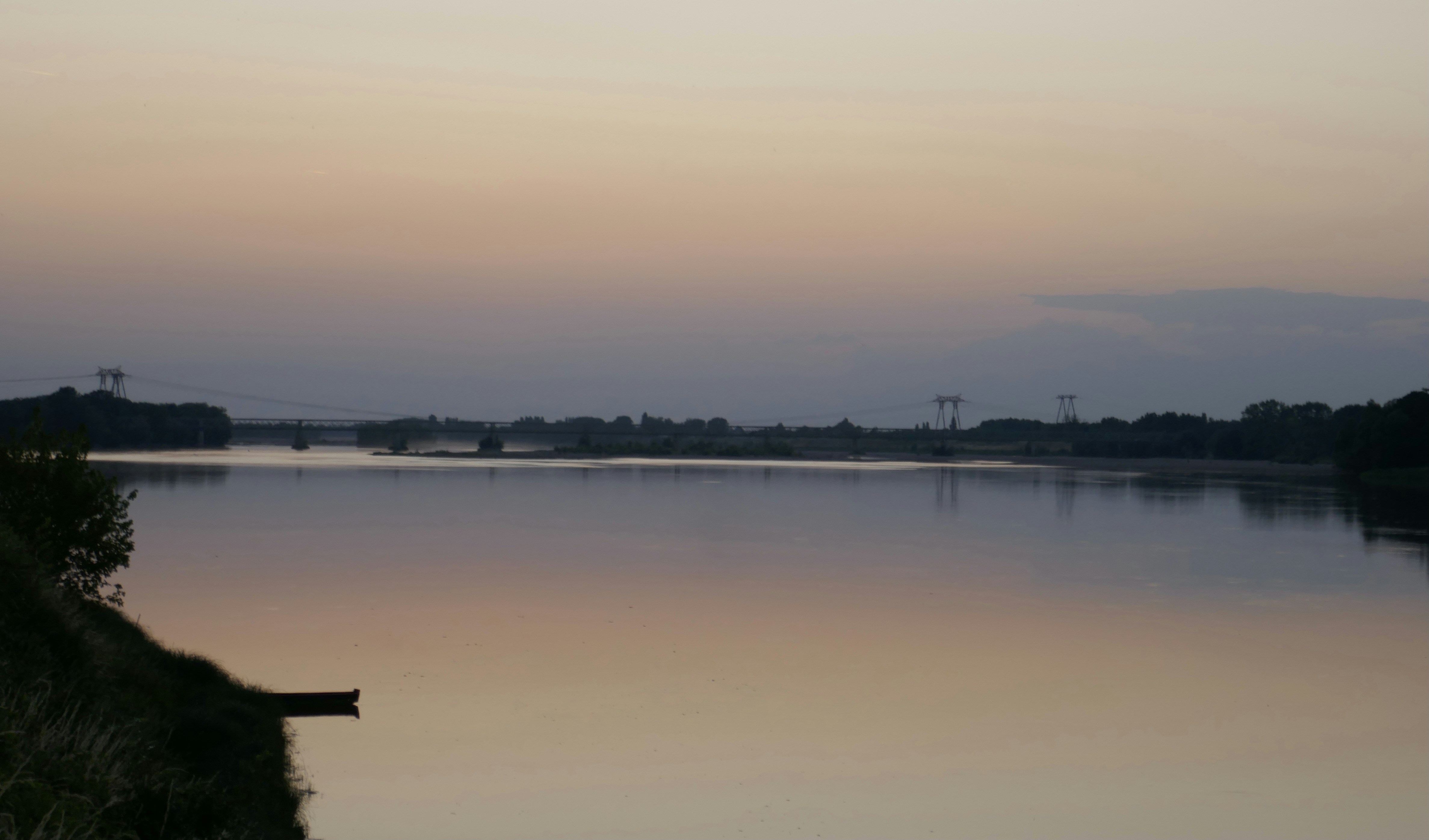 Calm river at twilight, reflecting soft hues of the sky with distant power lines and trees lining the shore.