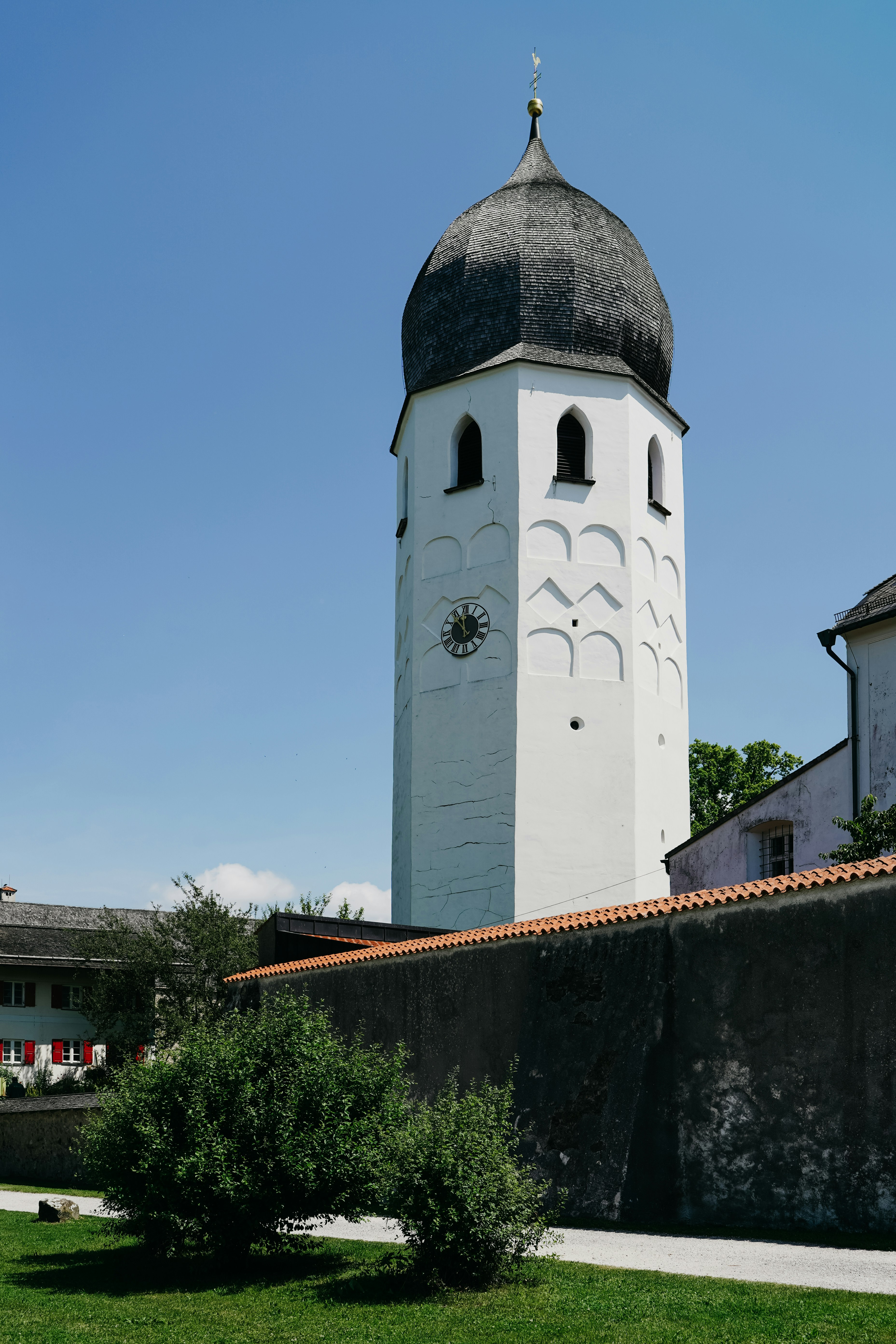 Historic white tower with a distinctive black dome, surrounded by lush greenery and a clear blue sky. The architecture reflects a blend of tradition and elegance.