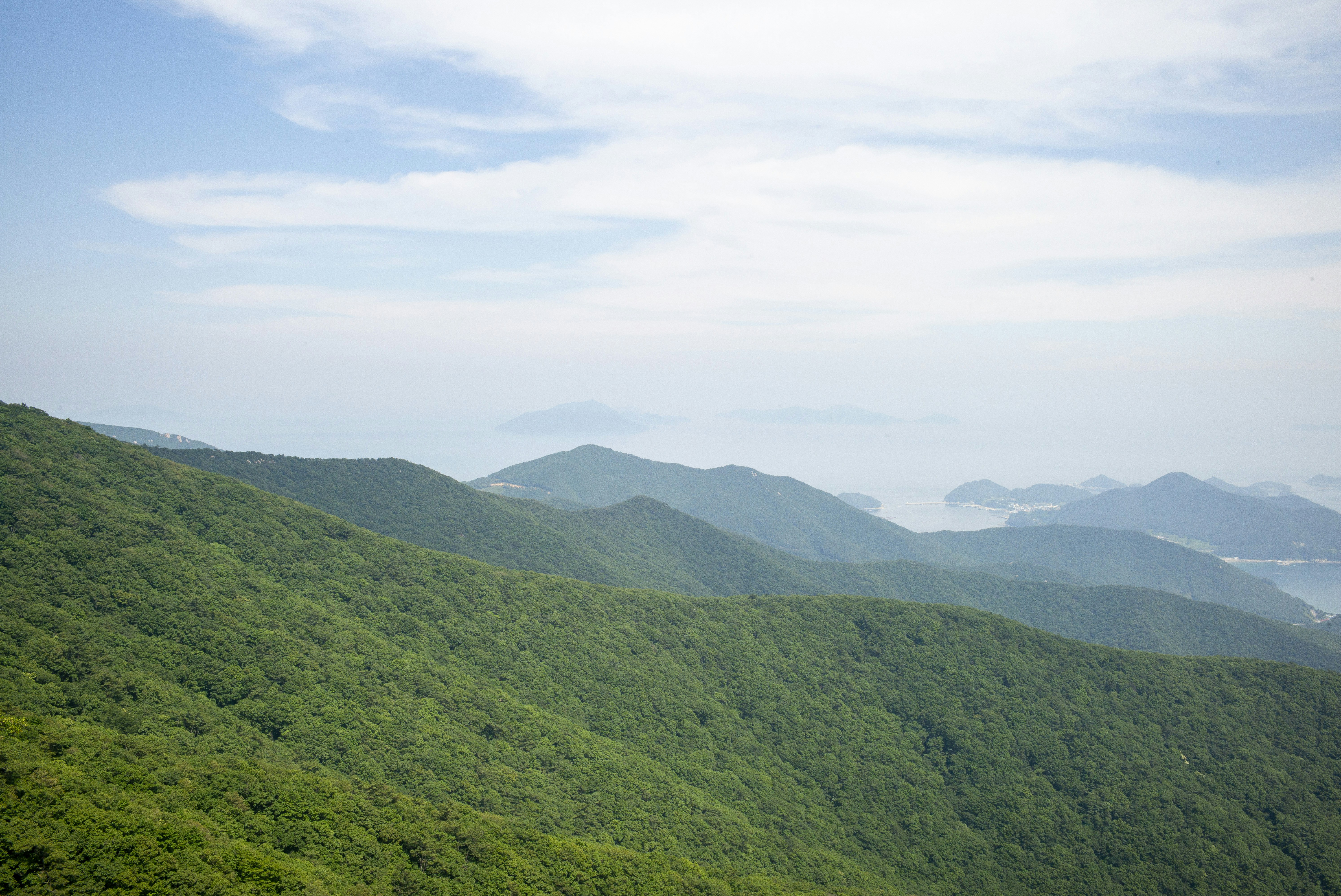 Lush green hills rolling towards the horizon, with distant islands barely visible in the misty sea. A serene landscape showcasing nature's tranquility.