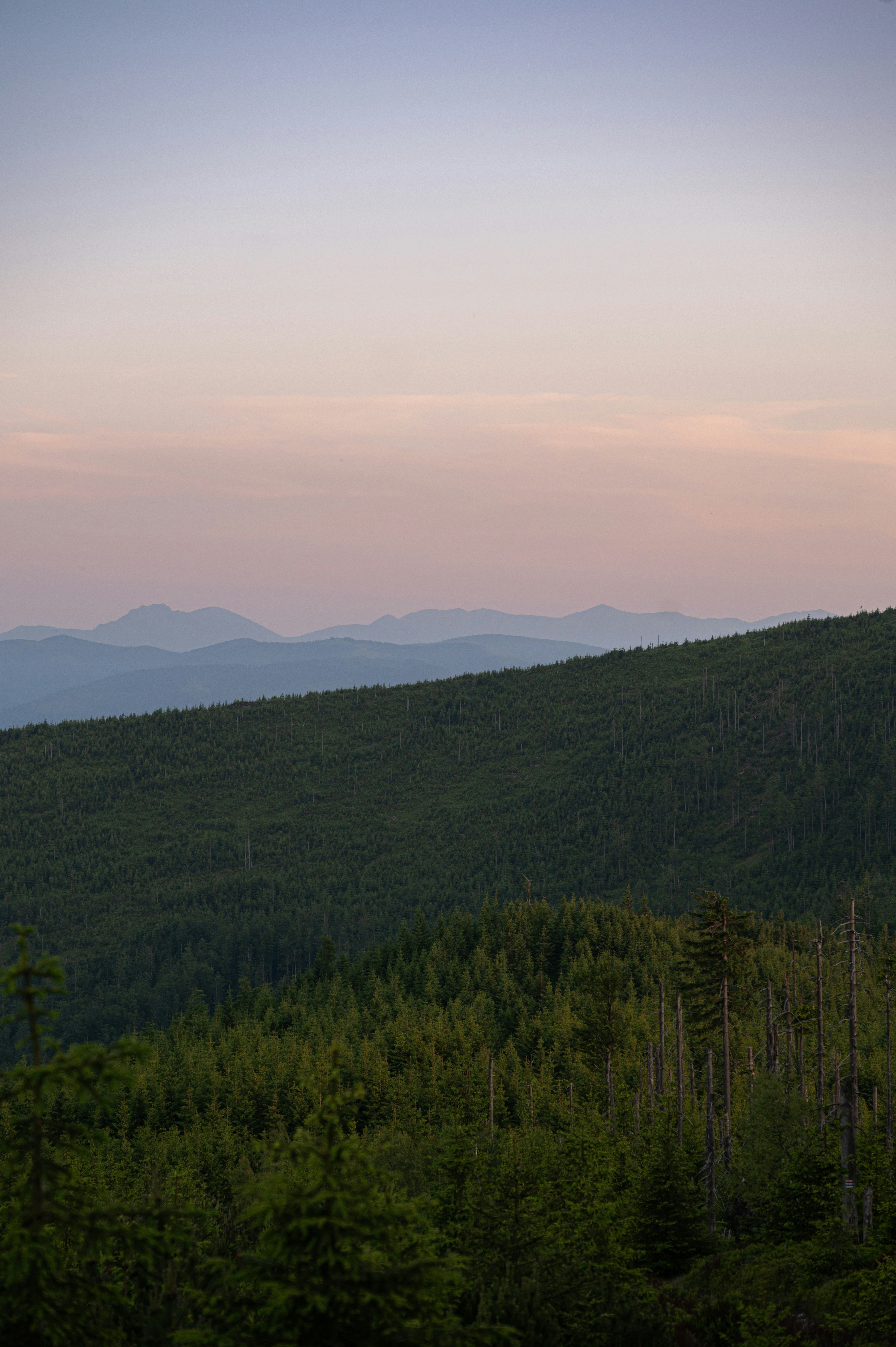 Lush green mountains stretch toward a soft, pastel sky at dusk, with faint outlines of distant peaks creating a serene landscape.
